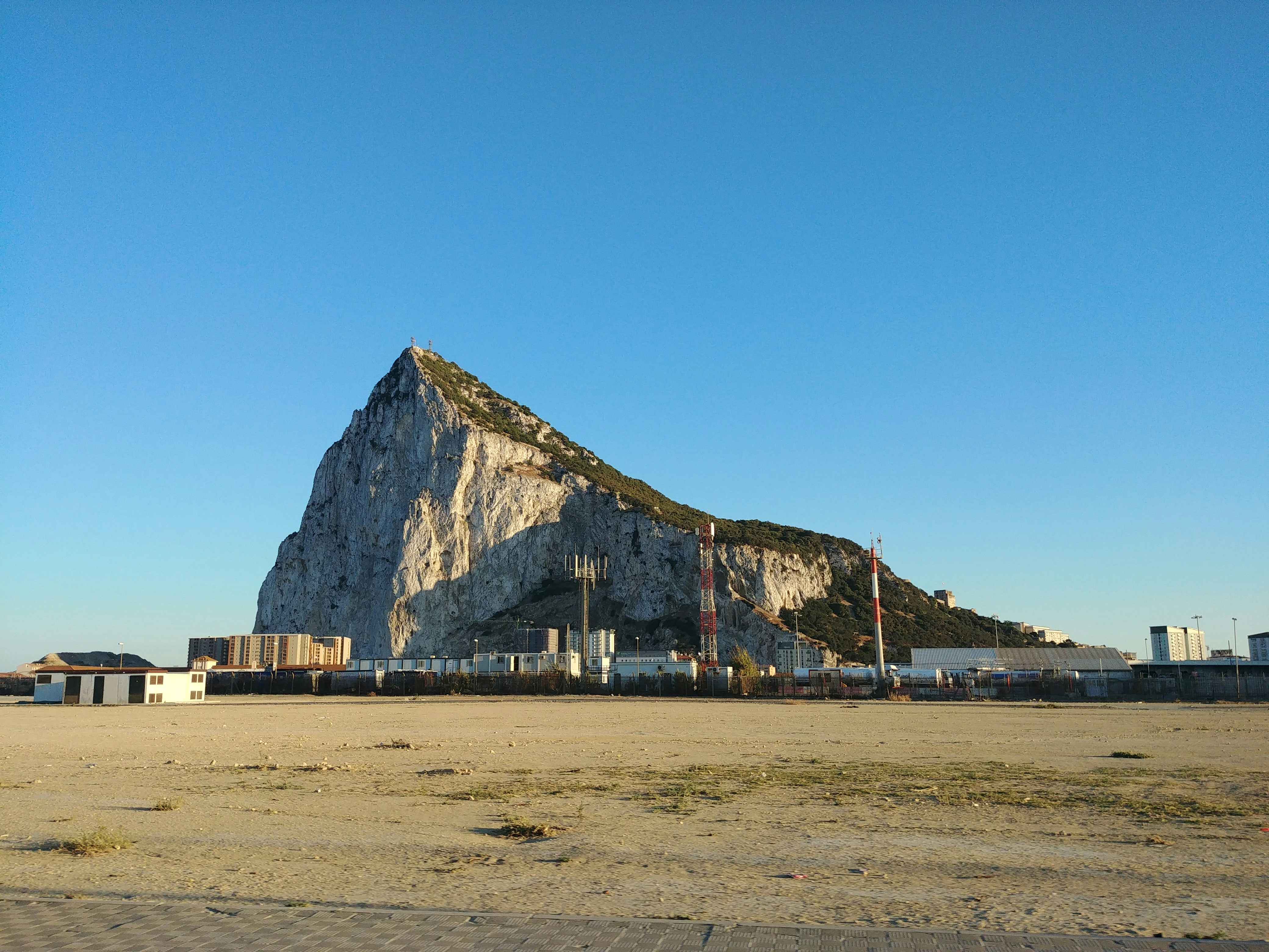 Massive coastal rock formation towers over a flat sandy shoreline and low-lying industrial promenade beneath a clear blue sky.