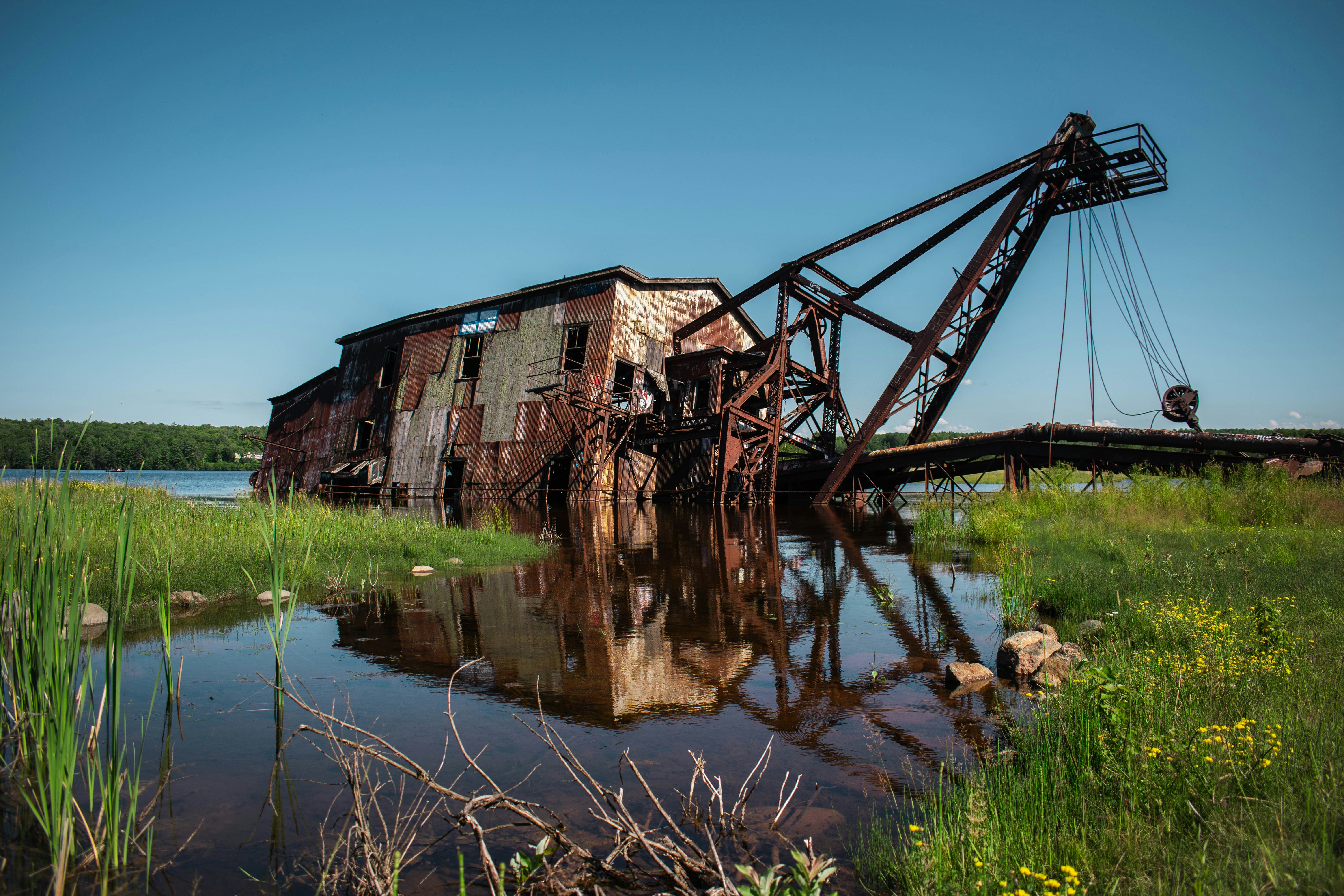 A dilapidated industrial structure partially submerged in water, surrounded by lush greenery and reflecting on the surface. The scene conveys a sense of abandonment and the passage of time.