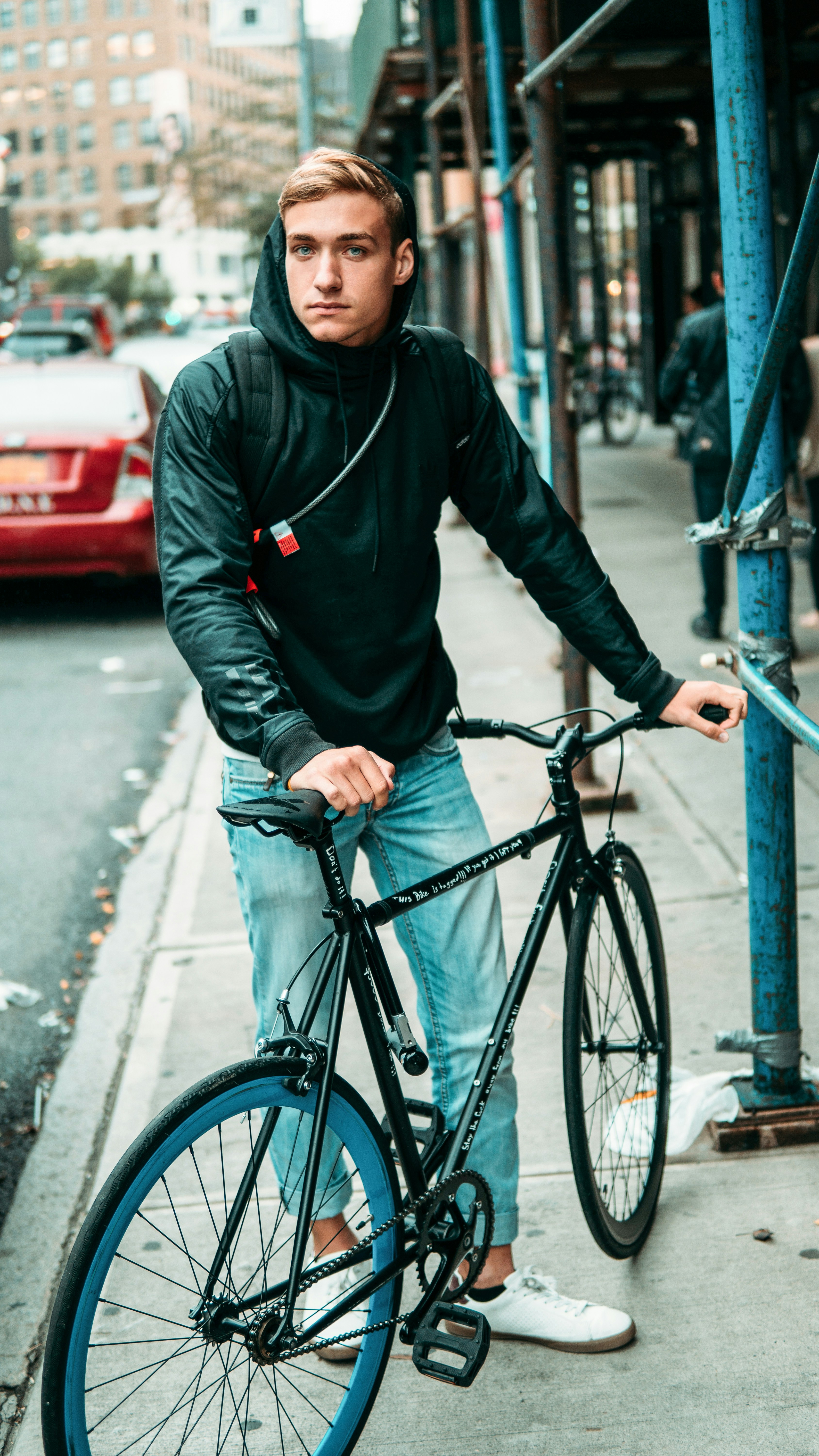 Young man in a black hoodie stands beside his bicycle on a city sidewalk, exuding a cool urban vibe.