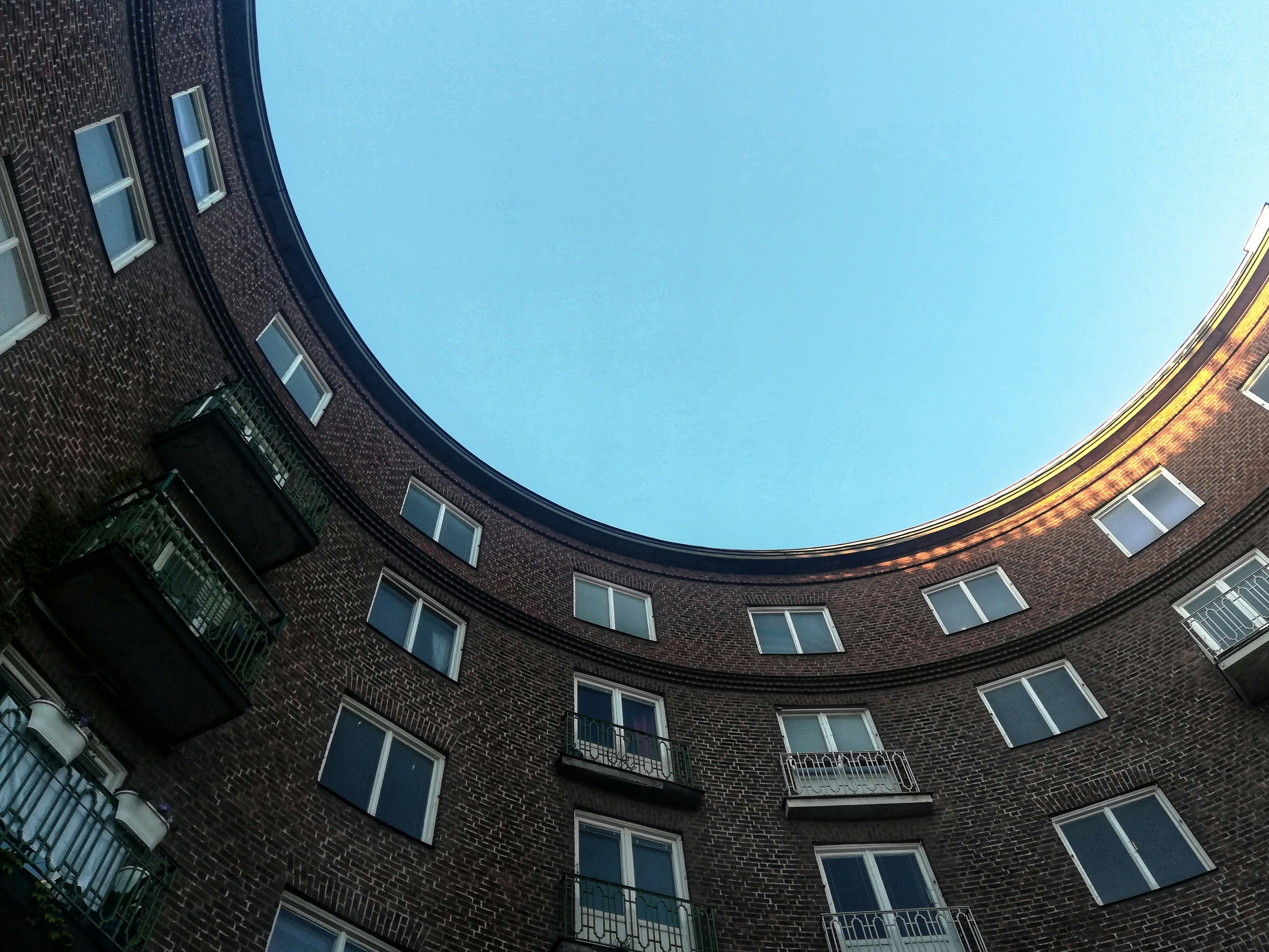 Architectural photograph of a circular brick courtyard with a clear blue sky opening framed by the walls. The composition emphasizes symmetry and depth in urban architecture.