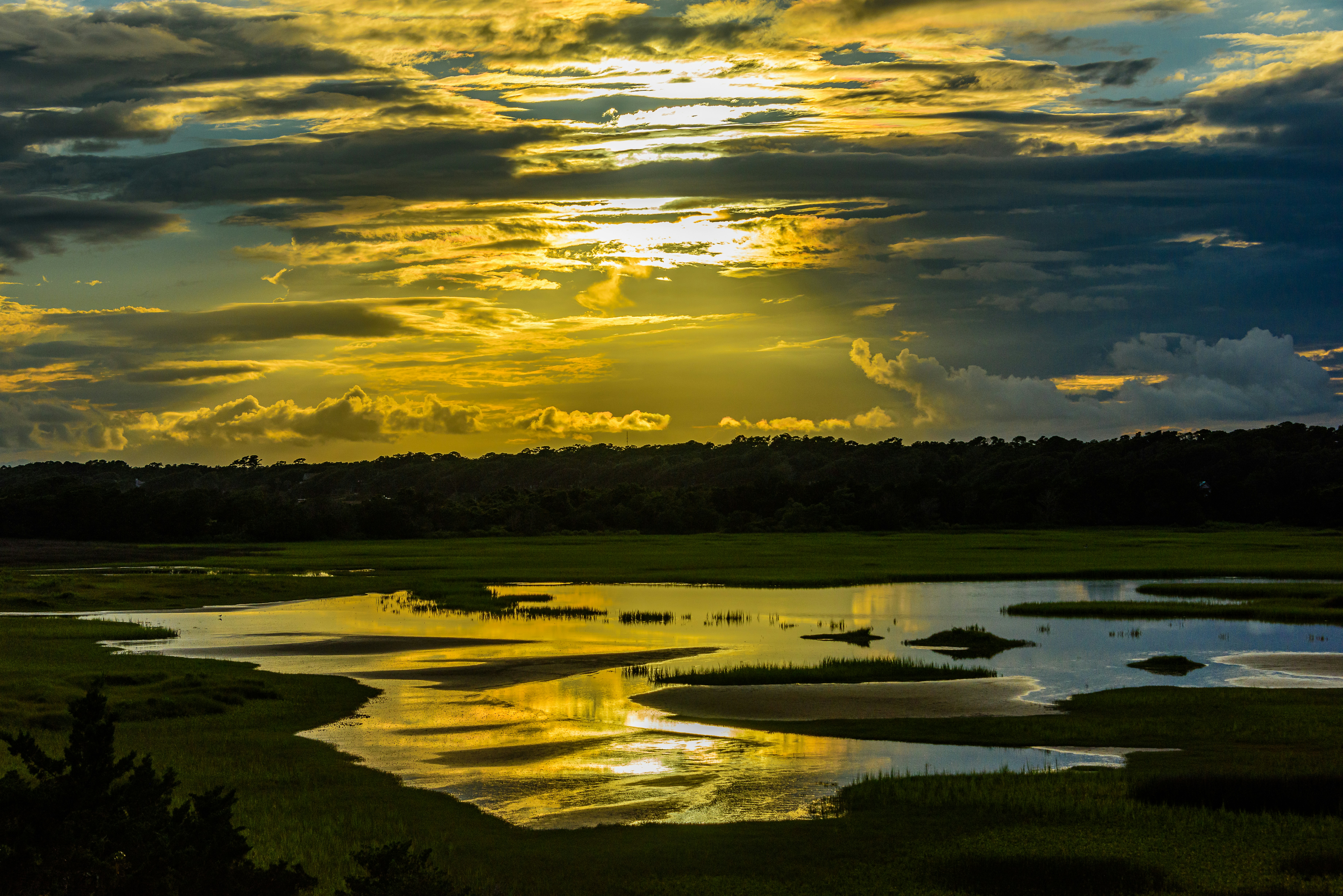 Golden sunlight reflects off tranquil marsh waters, highlighting the intricate patterns of the landscape under a dramatic sky.