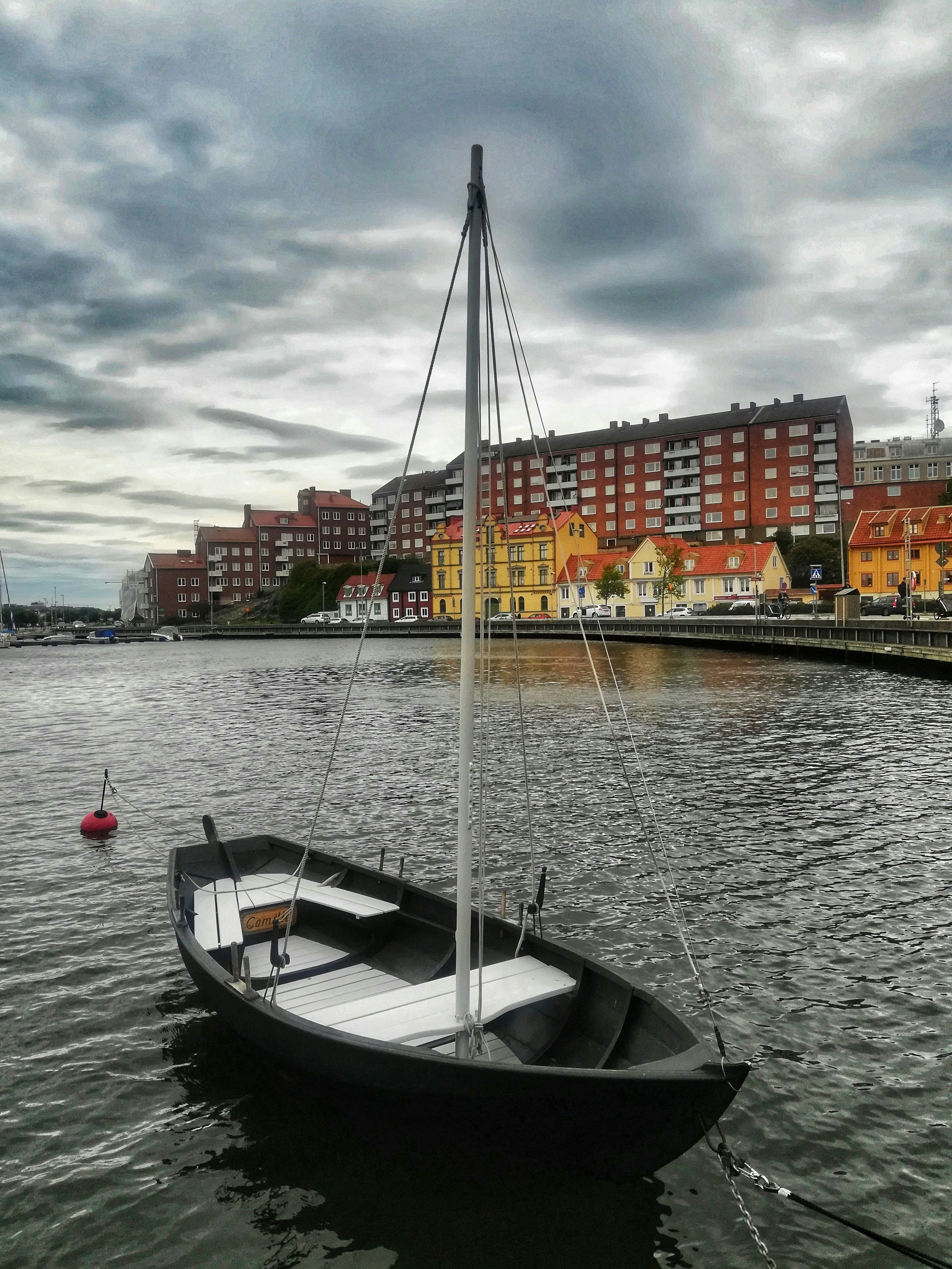 A black sailboat anchored in a tranquil harbor, with colorful buildings lining the waterfront under a cloudy sky.