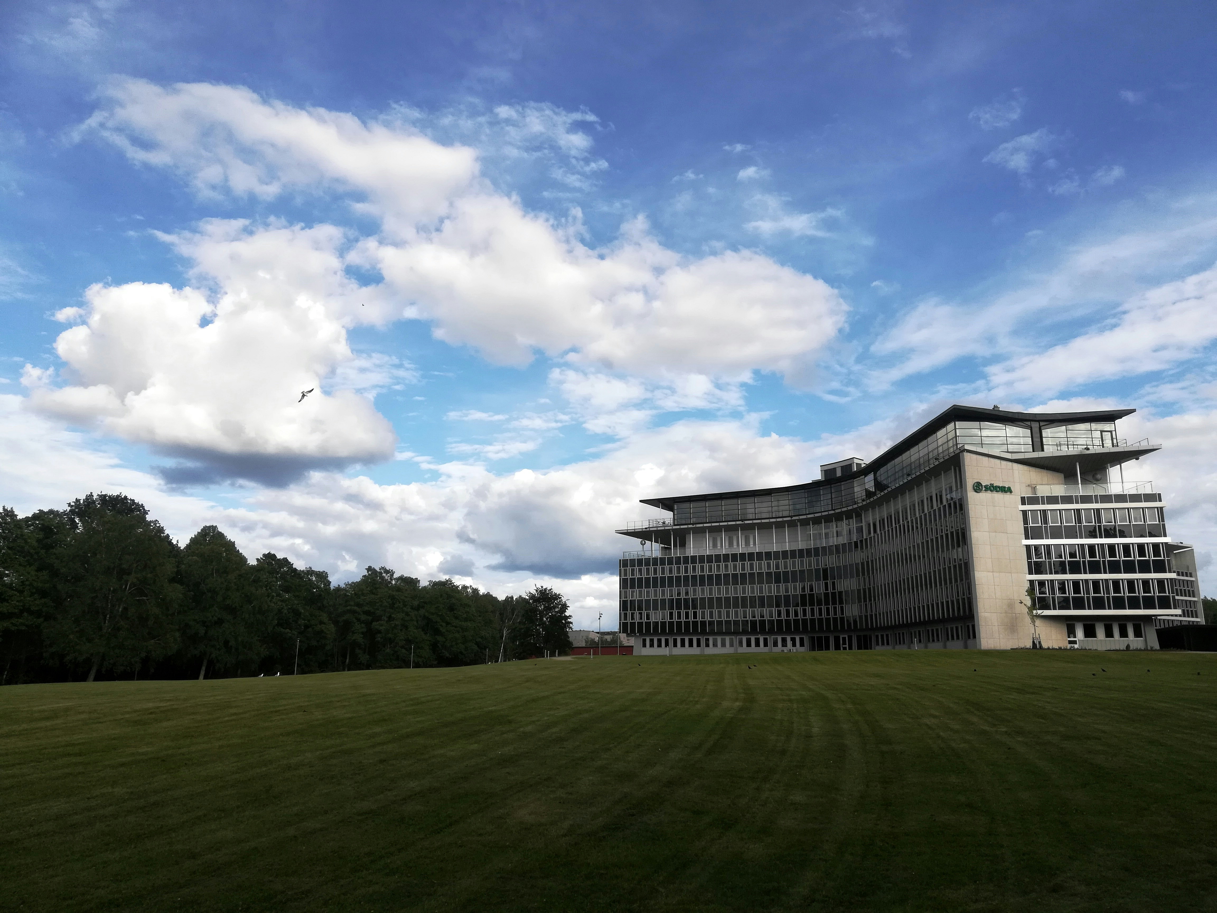 Contemporary building with large glass windows juxtaposed against a vibrant sky filled with fluffy clouds and greenery in the foreground.