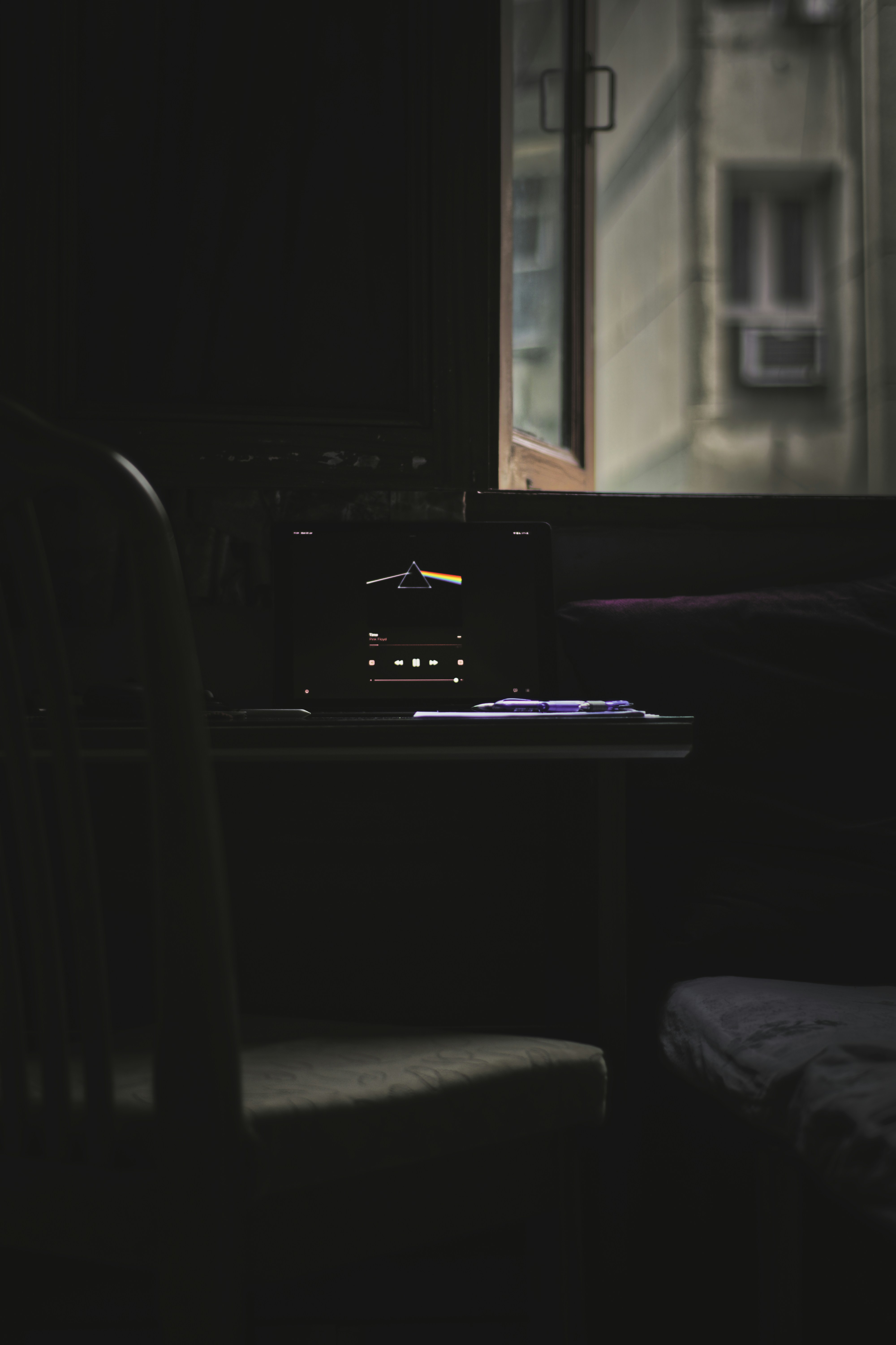 Dimly lit workspace featuring a laptop displaying a geometric interface, with a window revealing a muted urban backdrop.