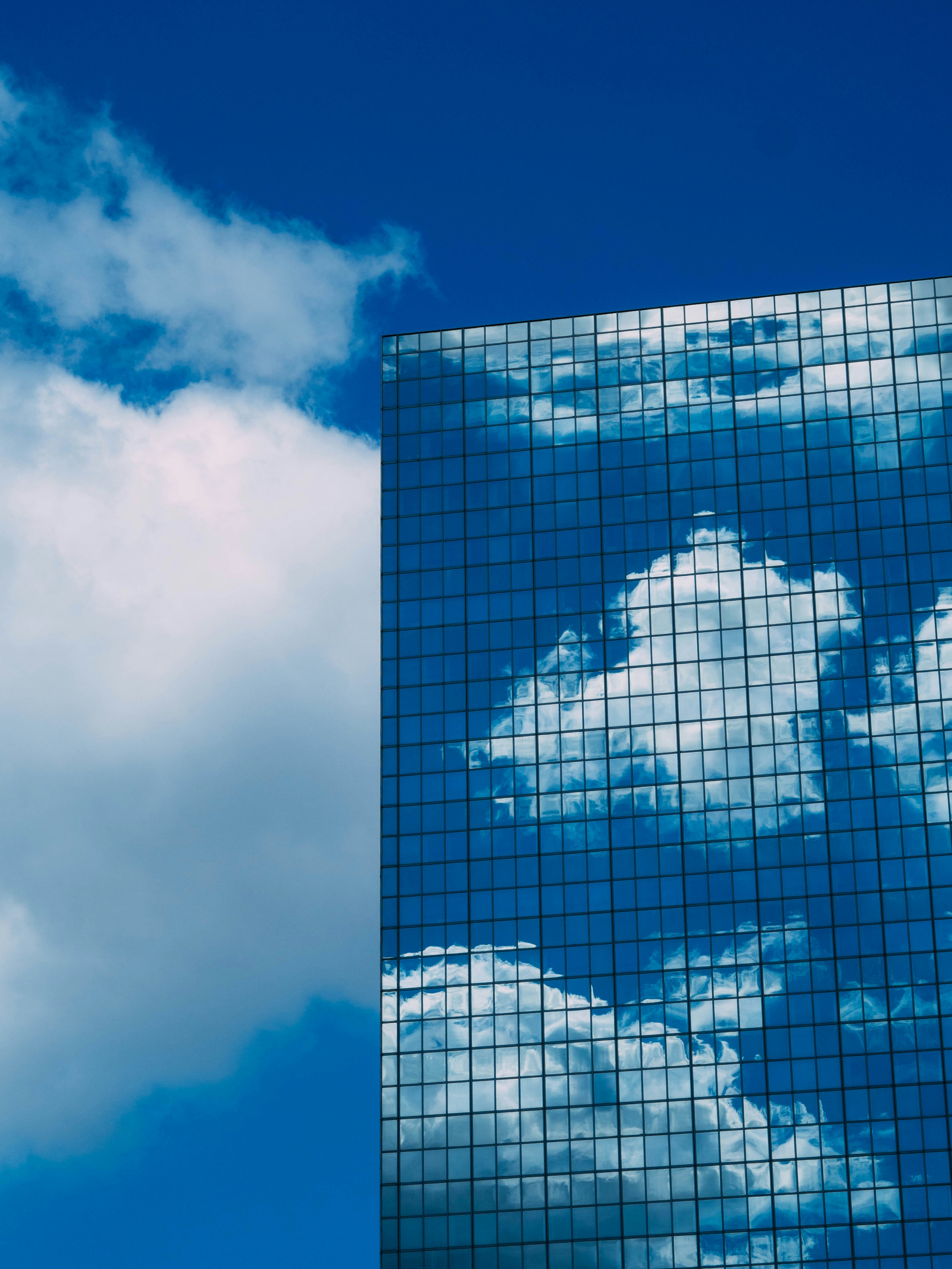 A glass building reflects fluffy white clouds against a vibrant blue sky, creating a harmonious blend of nature and architecture.