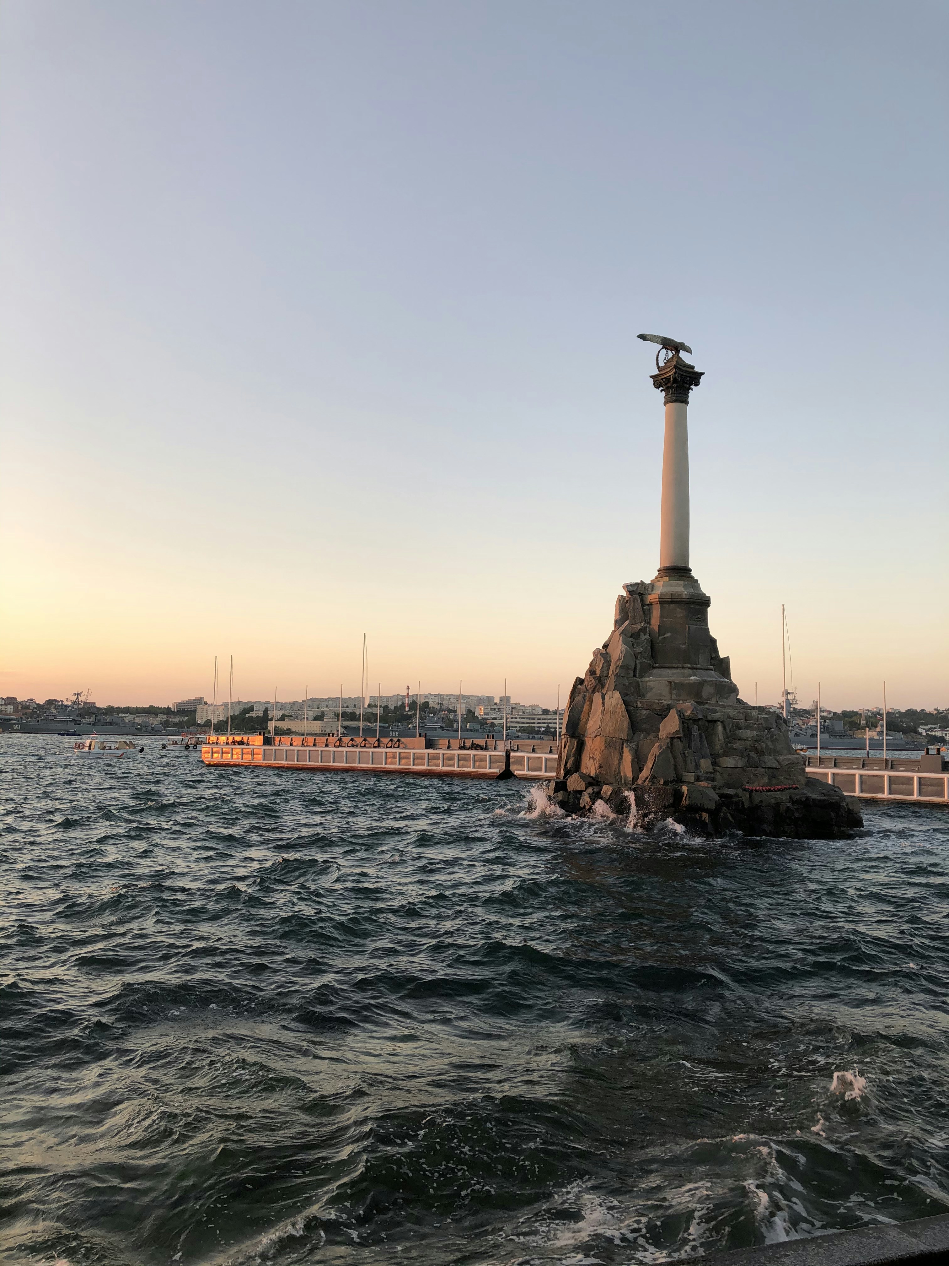 A monumental column topped with a statue stands amidst gentle waves at sunset, with a pier stretching into the distance.