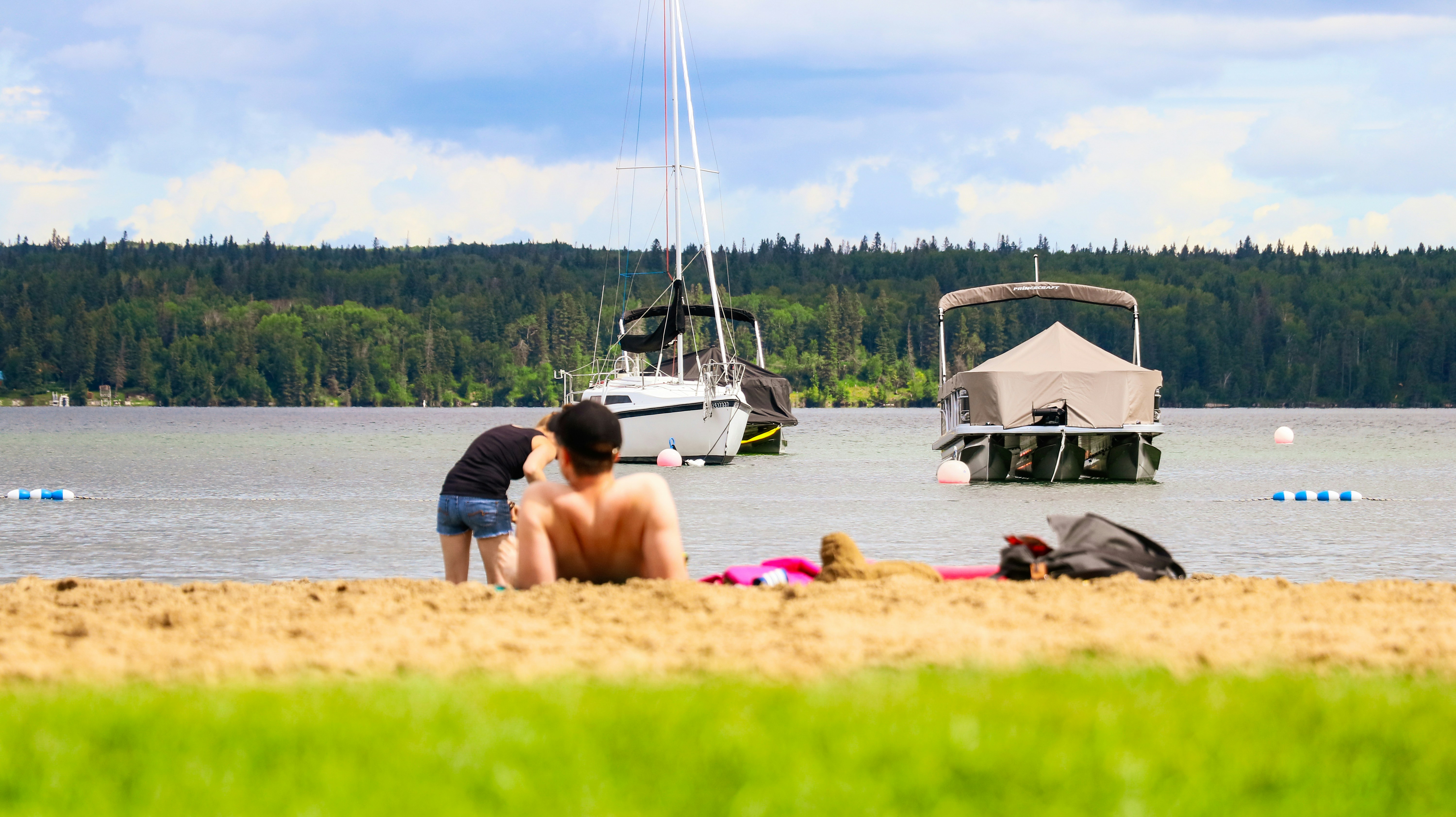 Two individuals enjoying a sunny day at the beach, with sailboats anchored in the calm waters behind them. A vibrant green lawn and sandy beach set the scene.
