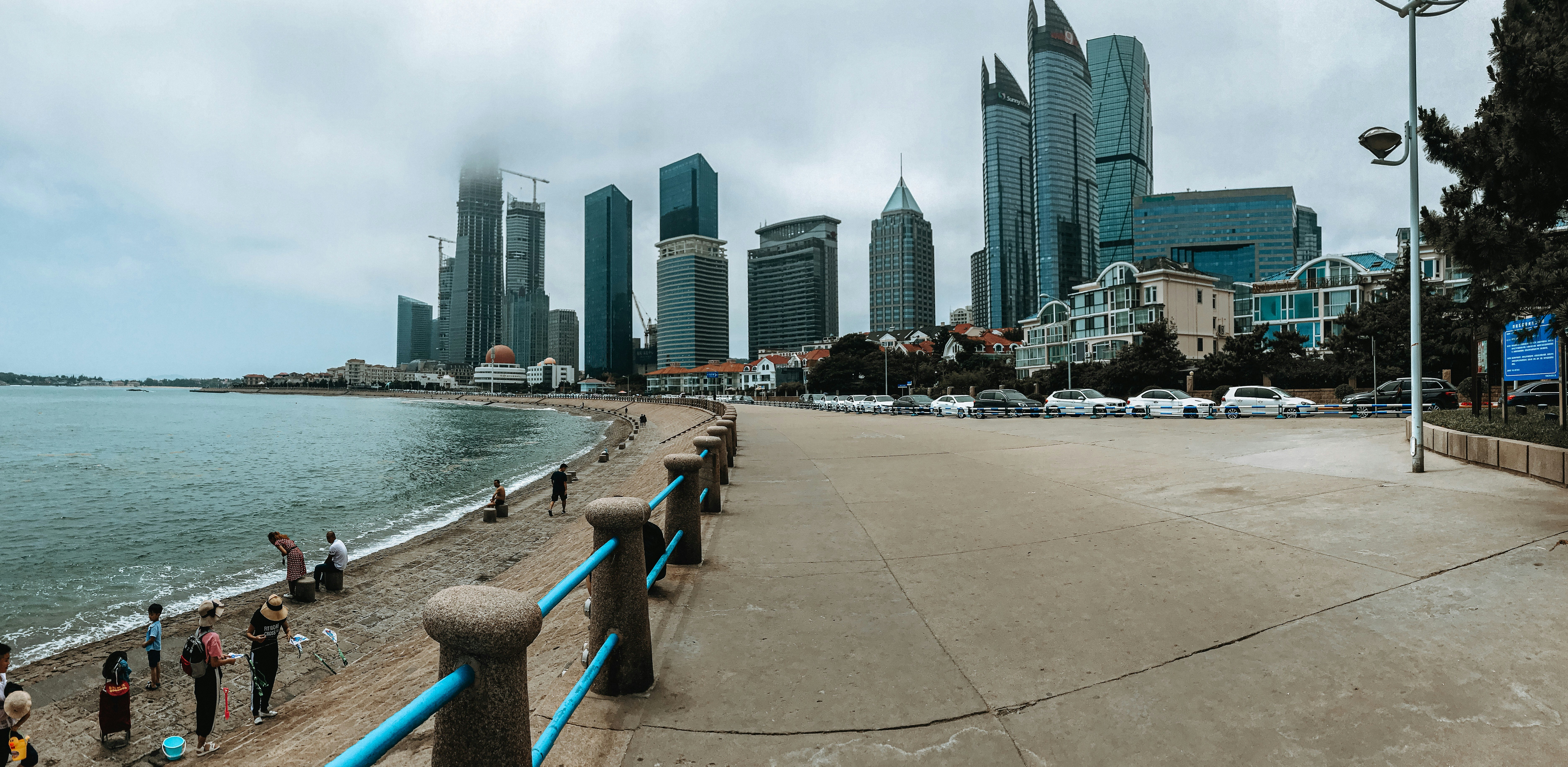 Modern skyscrapers line the waterfront promenade under a cloudy sky, with people walking along the beach.