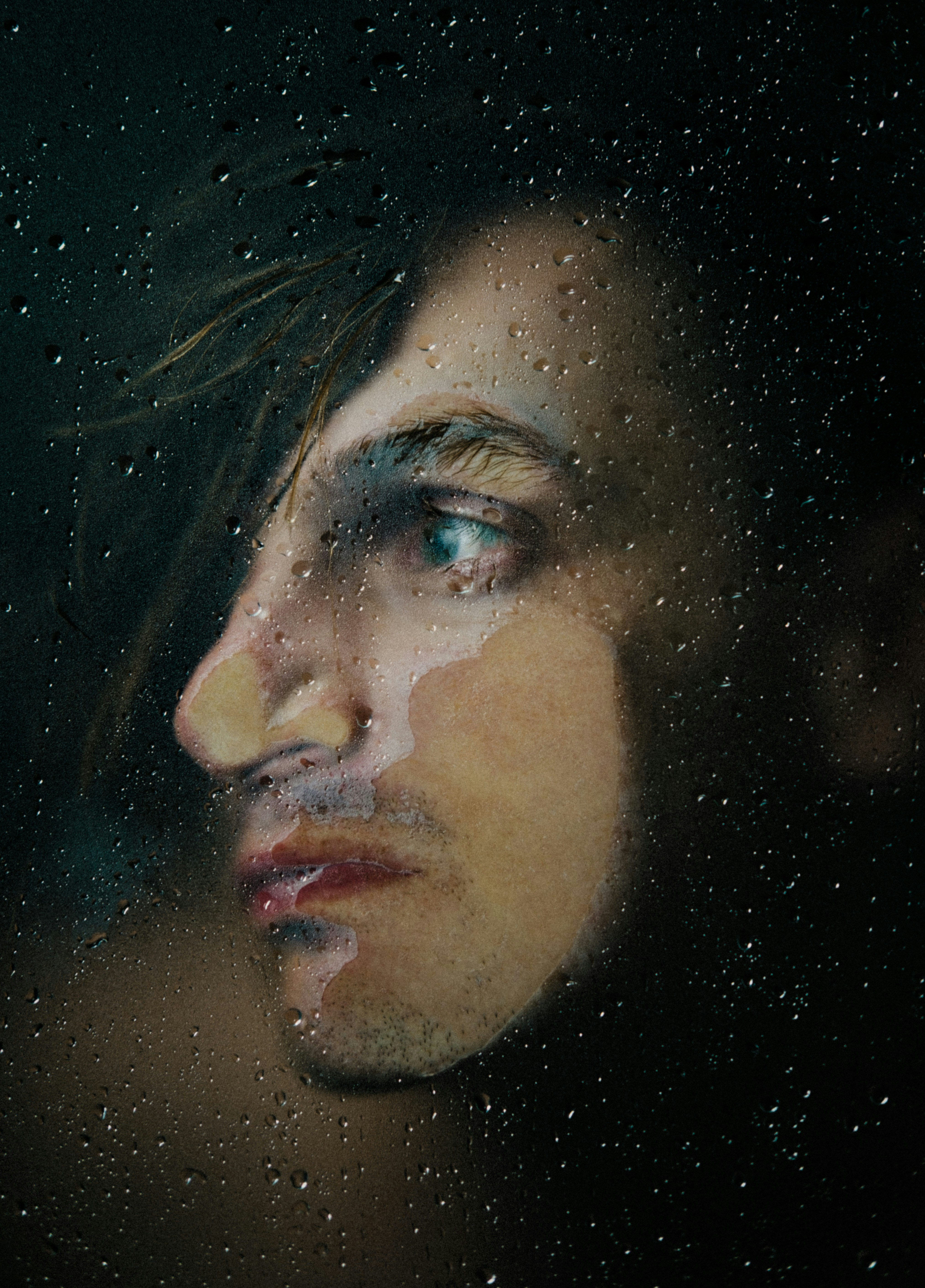 A close-up portrait of a person partially obscured by water droplets on glass, highlighting their contemplative expression.