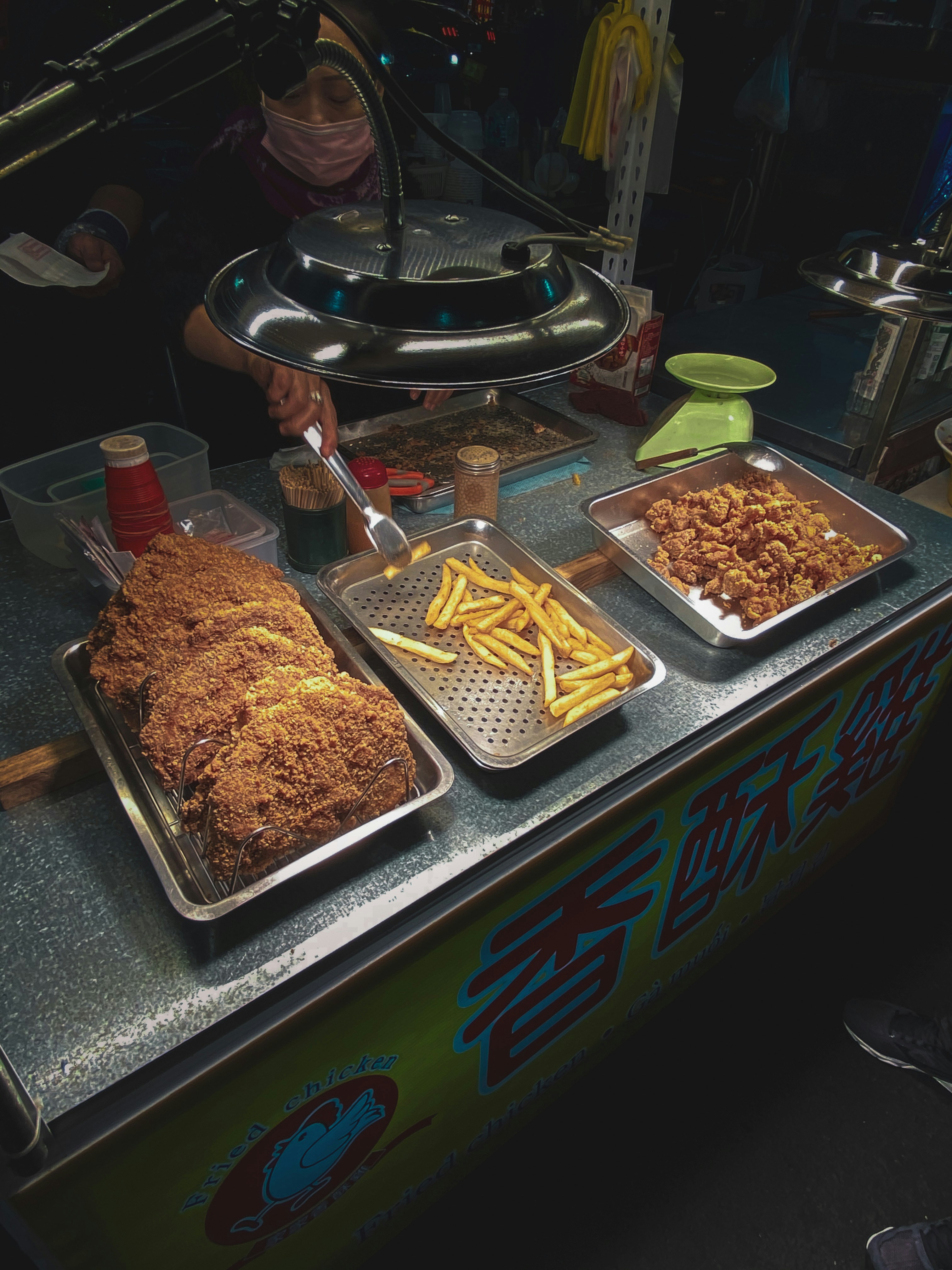 A vibrant food stall showcases crispy fried items, including chicken and fries, illuminated by a warm overhead lamp. The colorful signage adds to the lively atmosphere.