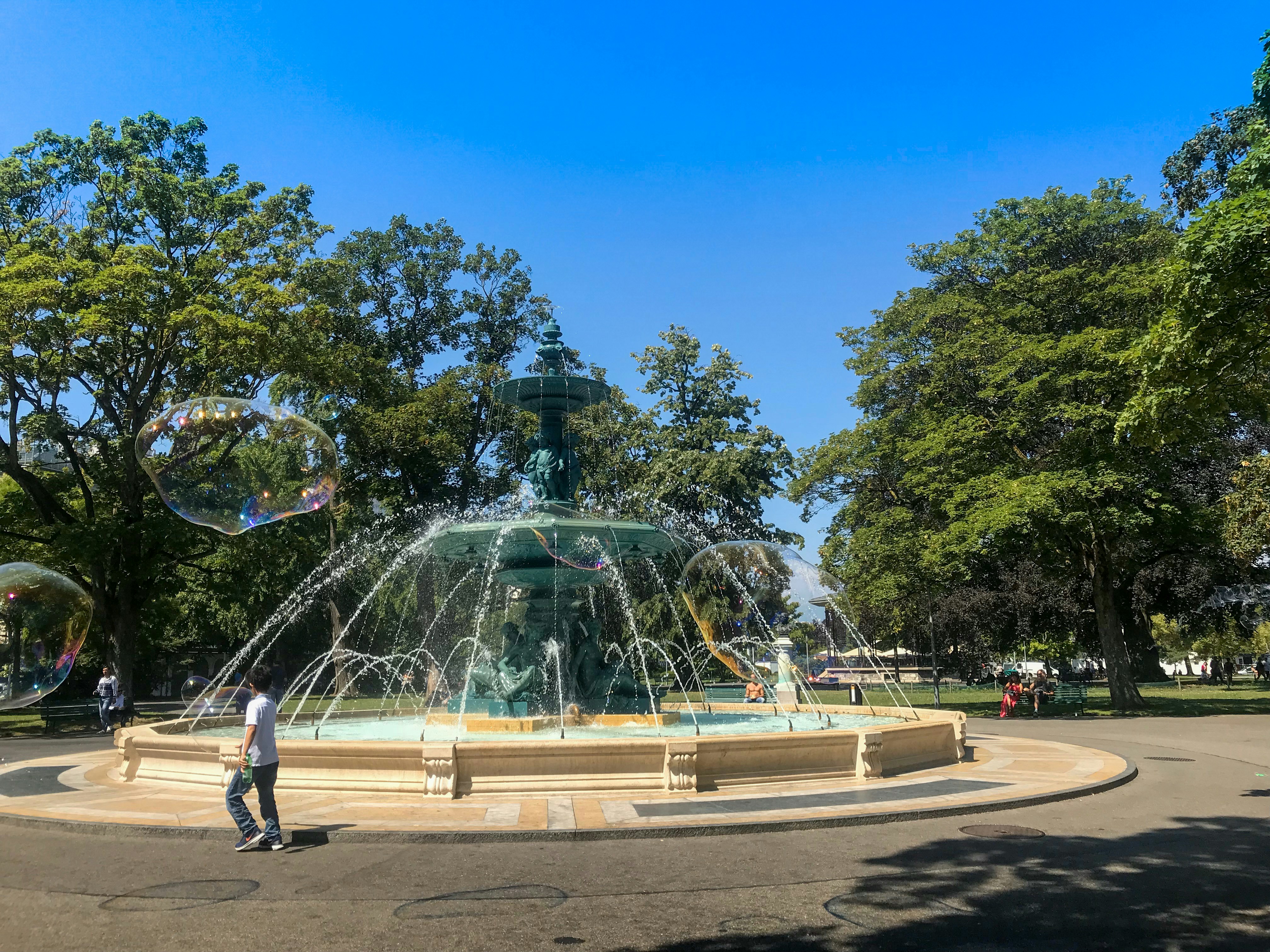 Ornate fountain encircled by lush trees under a clear blue sky.