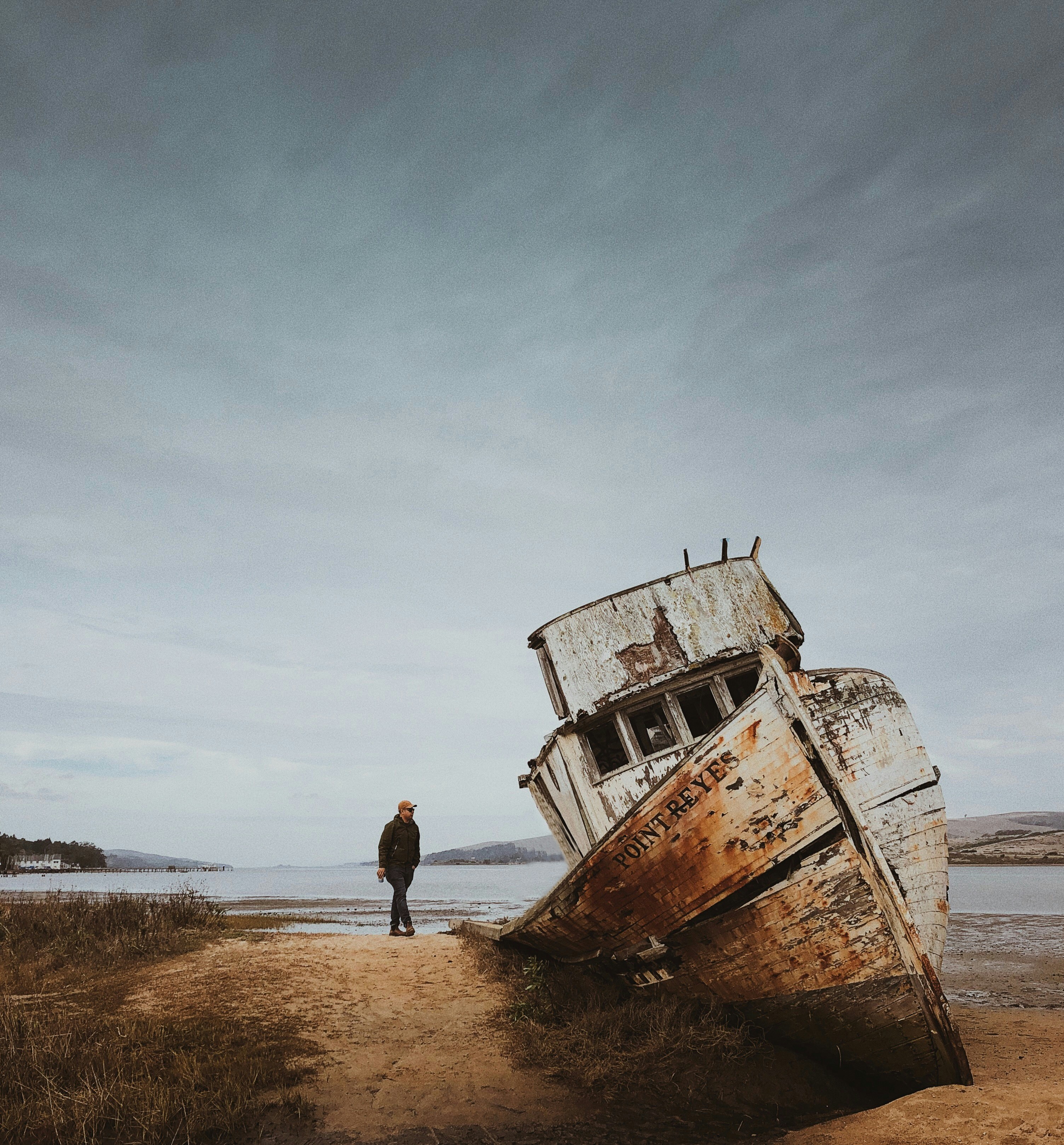 Weathered fishing boat resting on a sandy shore, with a solitary figure walking nearby. The scene evokes a sense of nostalgia and abandonment.