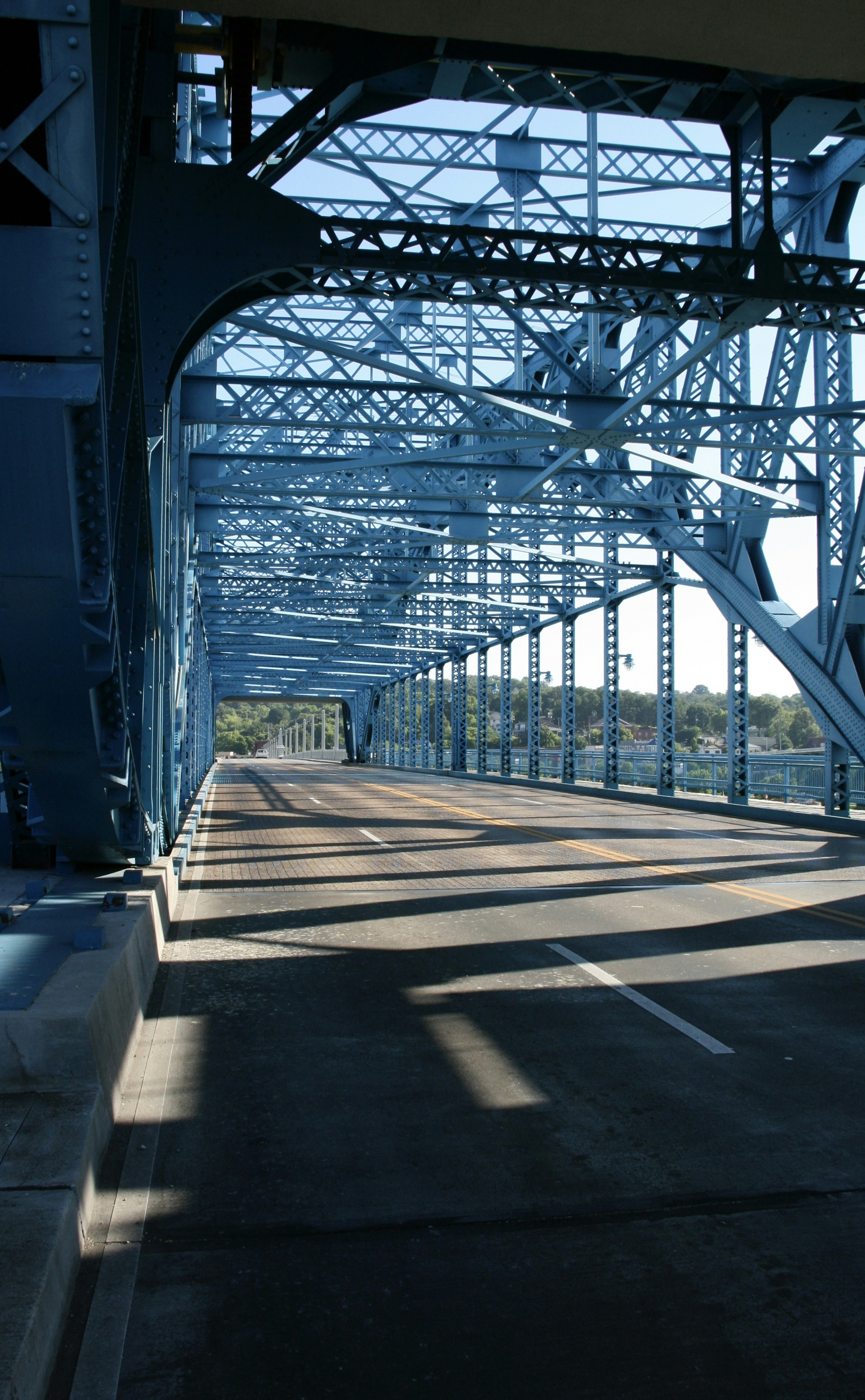 Intricate blue steel framework of a bridge with a wide, empty roadway beneath a clear sky. Shadows cast across the surface enhance the structural design.
