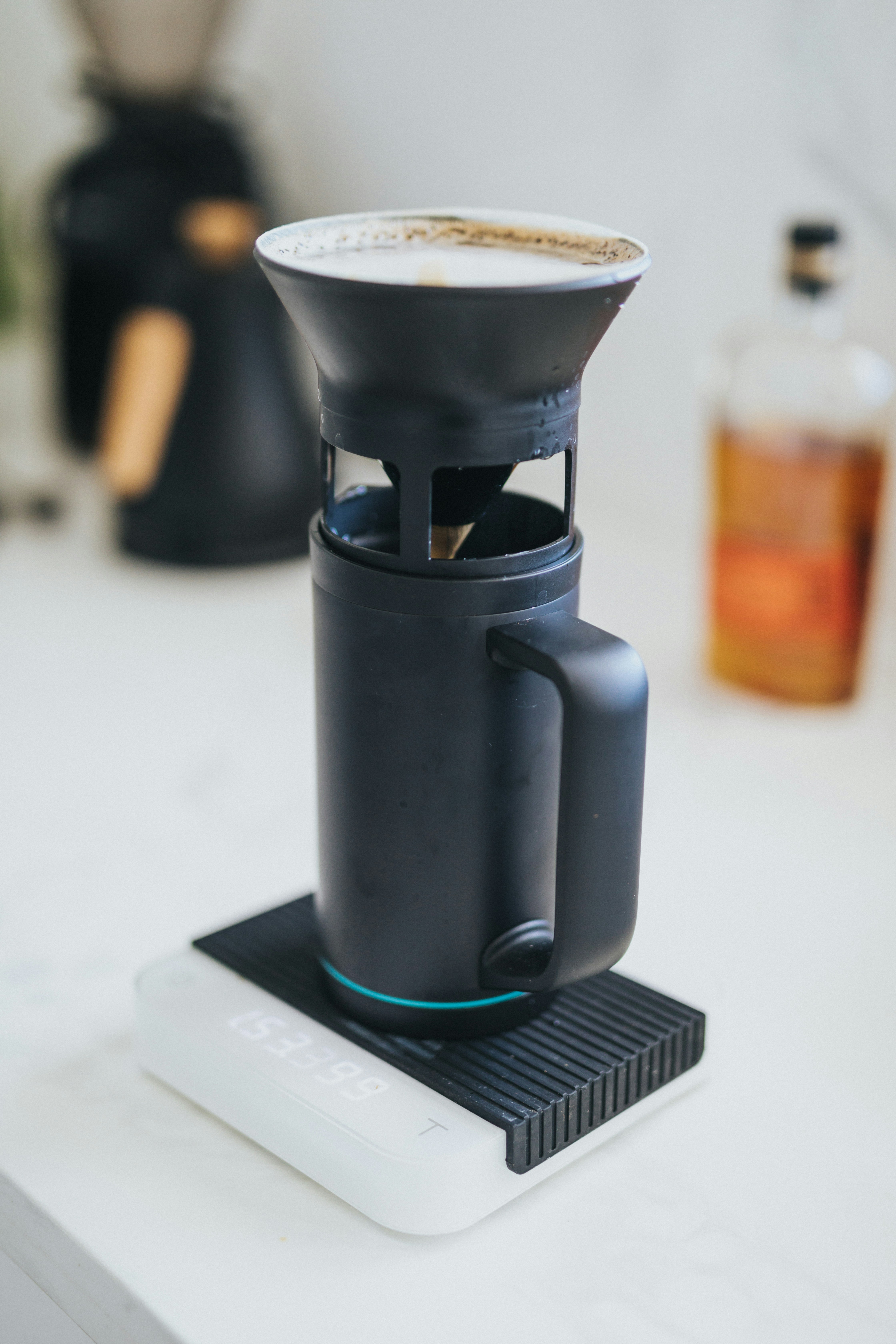 A sleek black coffee dripper sits atop a digital scale, with freshly brewed coffee filtering through. In the background, a warm bottle adds to the cozy atmosphere.
