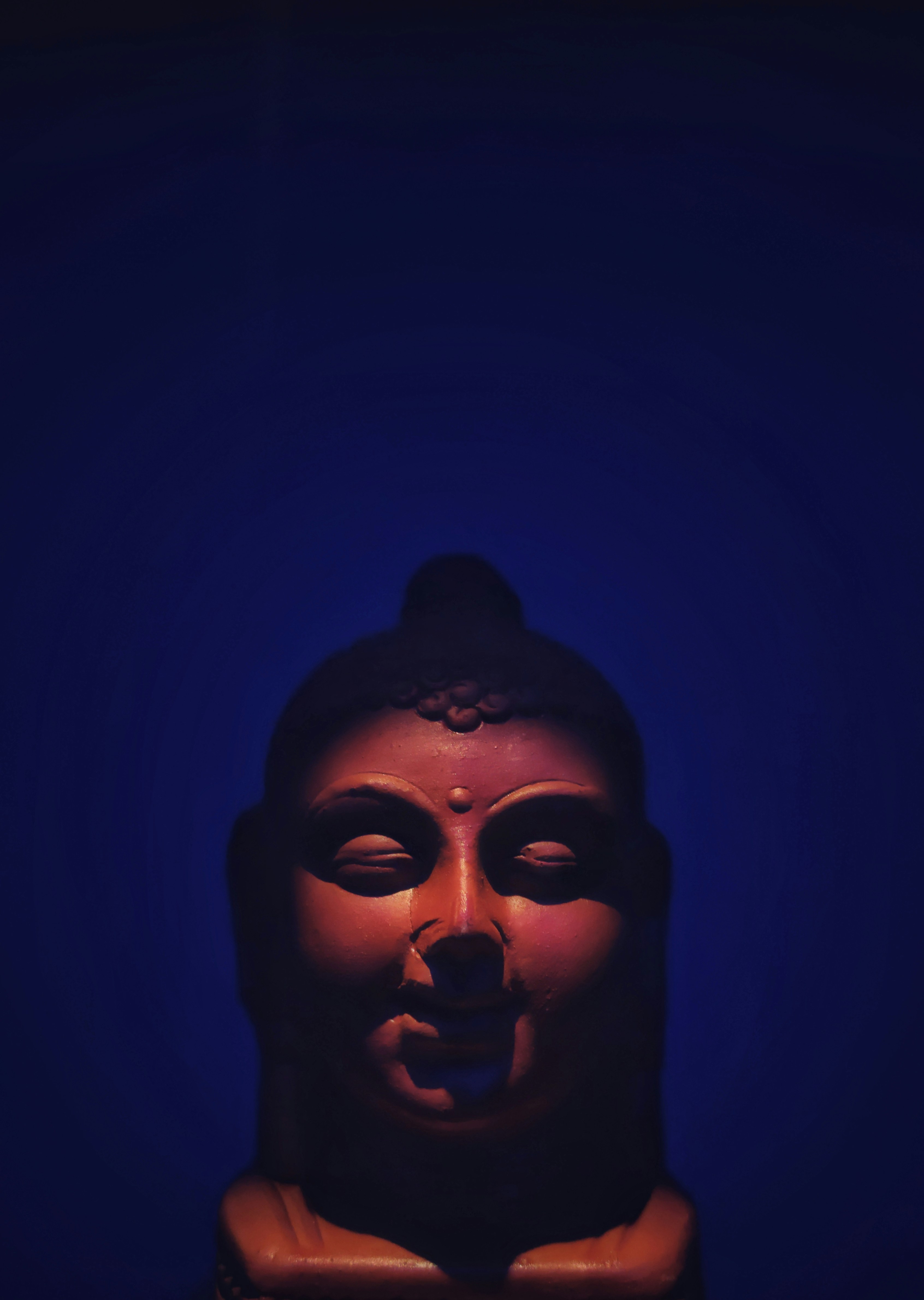 Close-up of a Buddha bust sculpture bathed in deep blue light against a dark background. The serene expression is highlighted by soft, directional illumination.