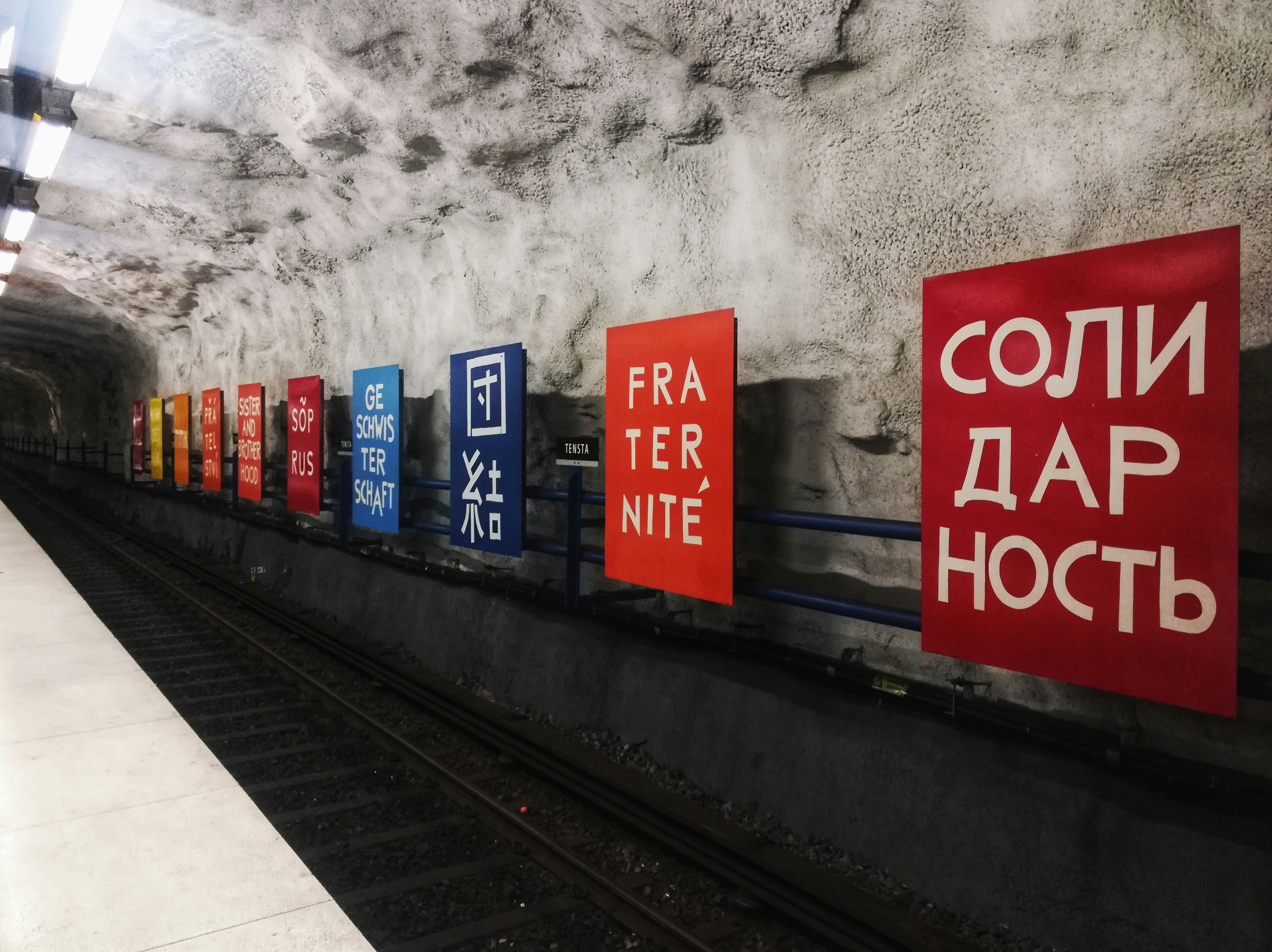 Colorful signs featuring various languages promoting unity and friendship line a subway platform. The artwork creates a vibrant atmosphere in an urban setting.