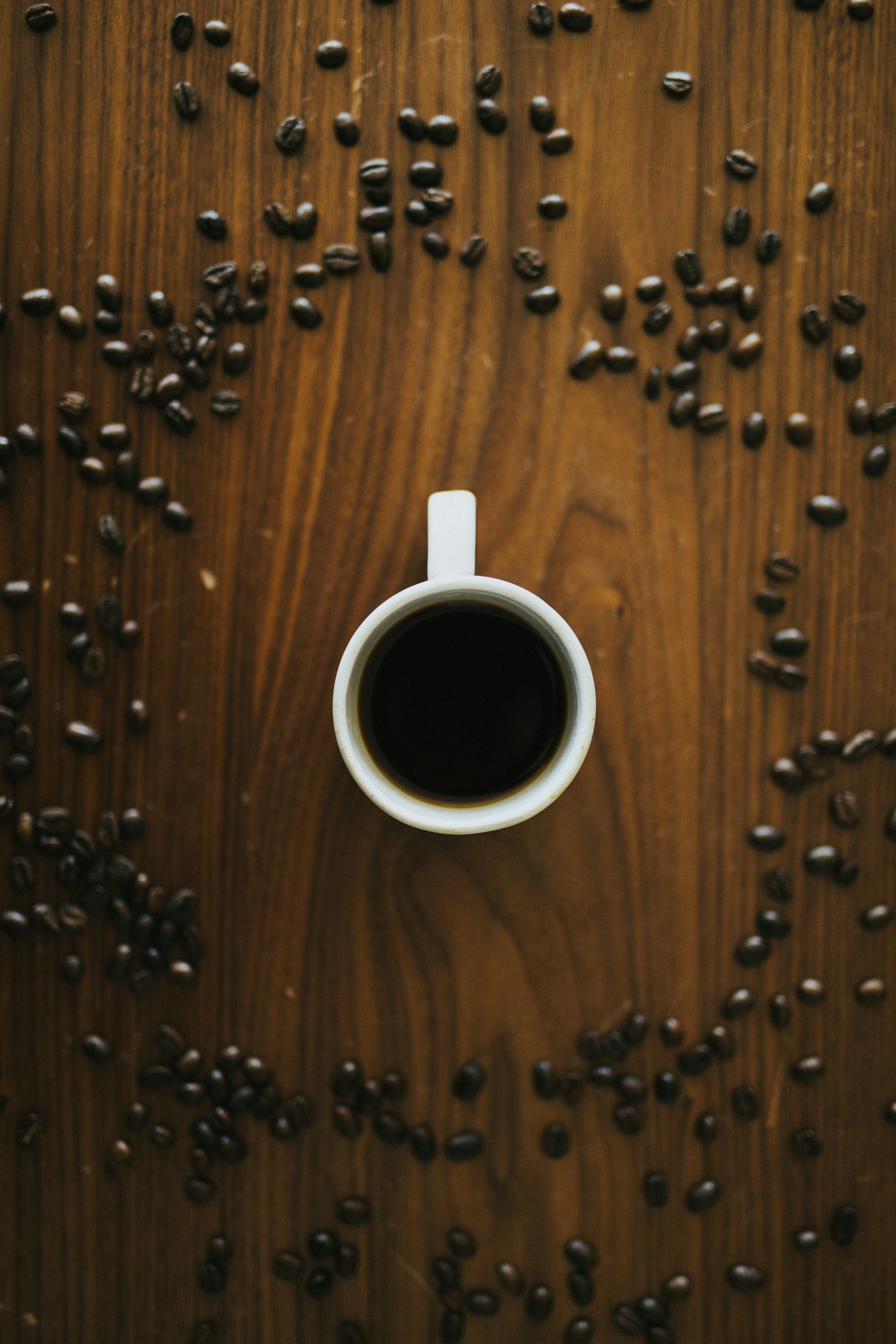 A white coffee cup filled with dark coffee sits atop a wooden table, surrounded by scattered coffee beans, creating a warm and inviting atmosphere.
