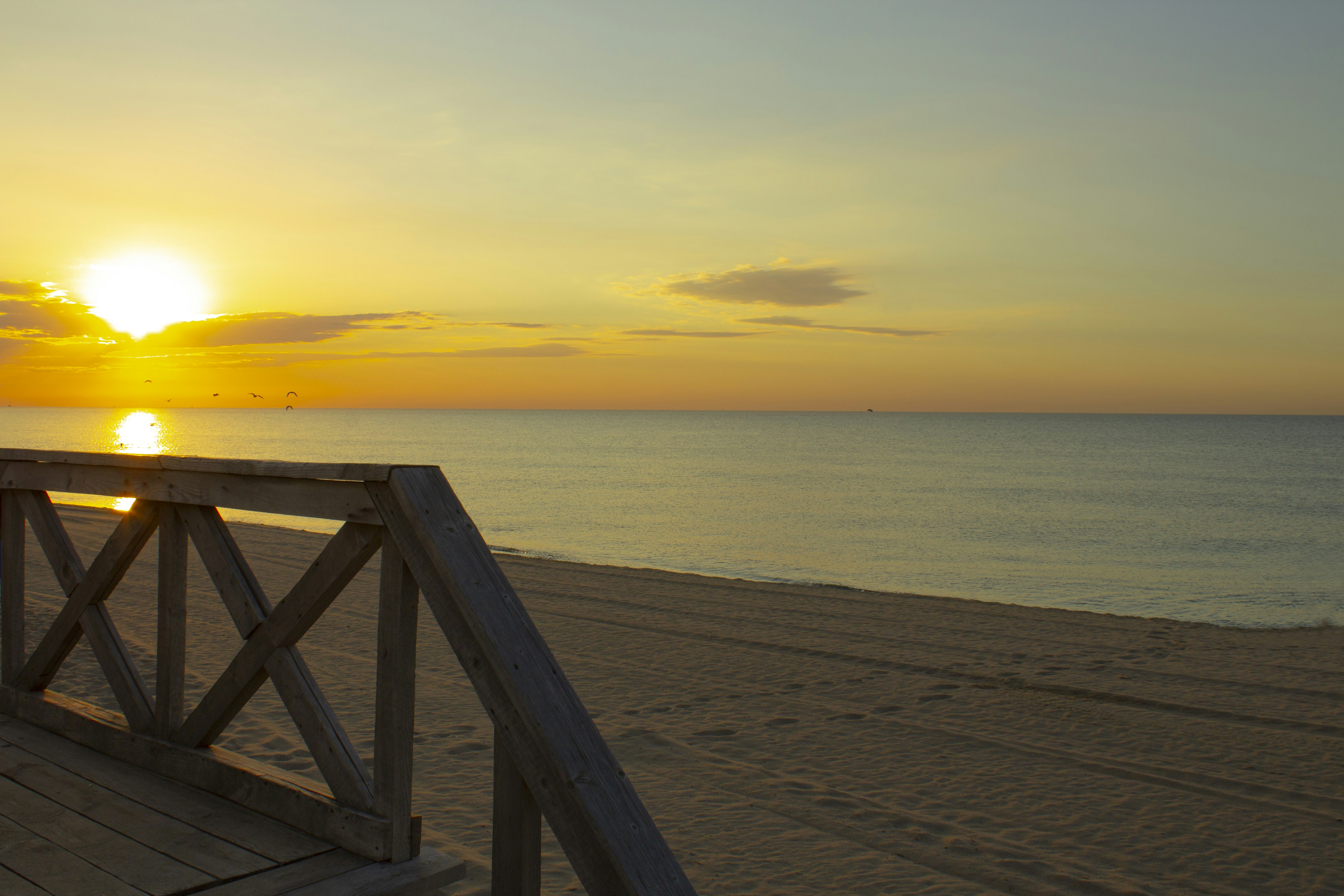 Sunset casting golden hues over calm ocean waters, framed by a wooden railing on the beach.