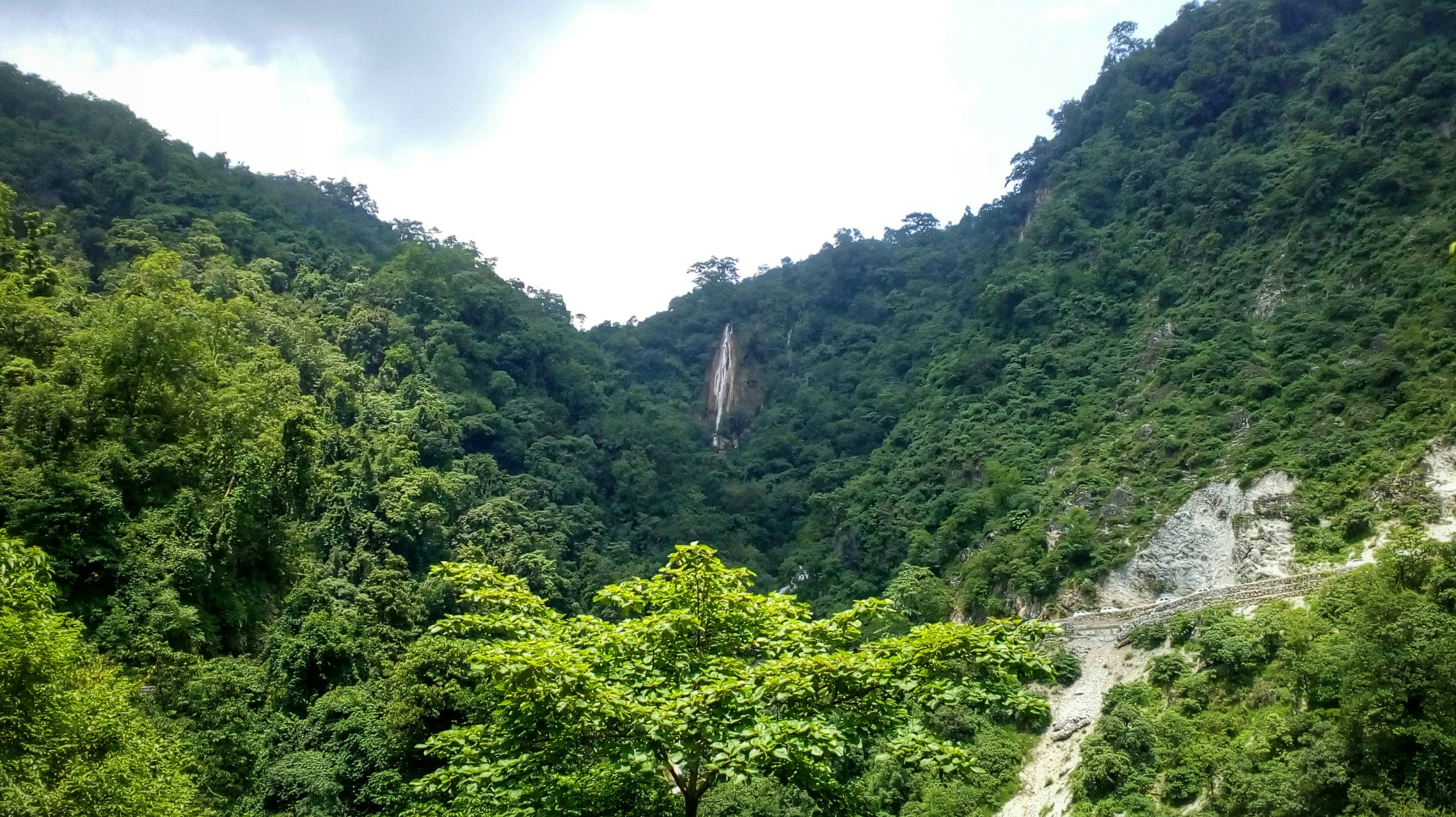A serene landscape featuring a waterfall cascading down lush green mountains, framed by vibrant foliage and rocky terrain.