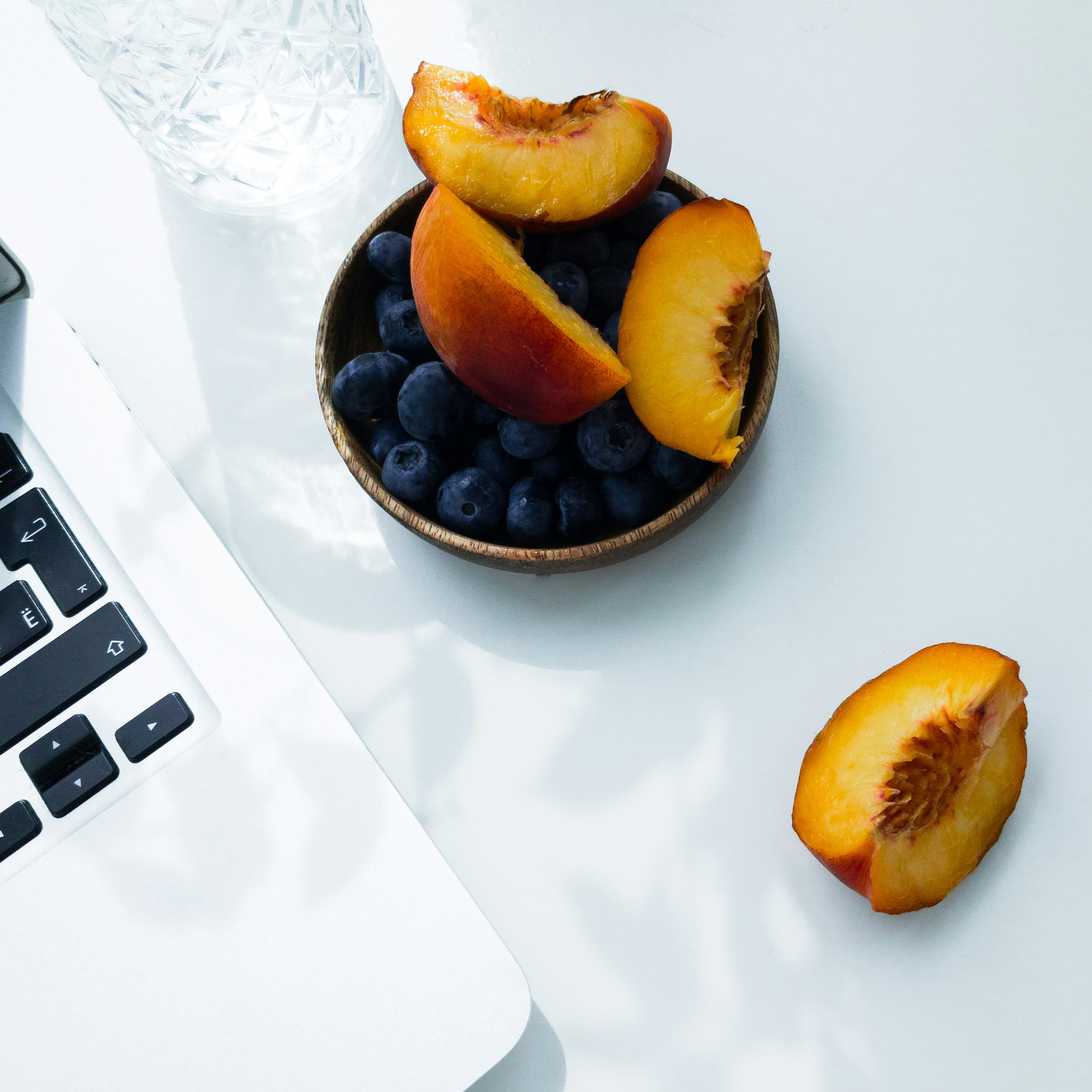 Sunlit Bowl of Blueberries and Peach Slices Beside a Laptop on a White Desk