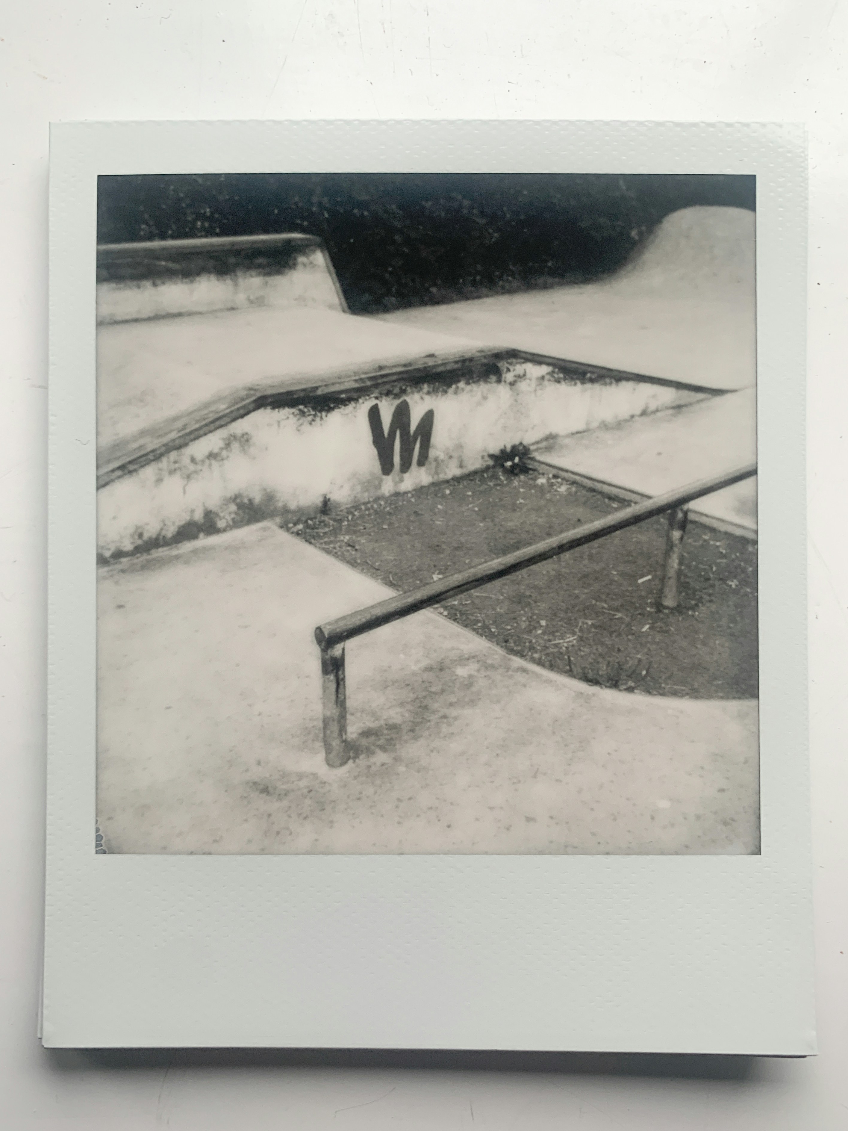 A monochrome view of a skatepark featuring a graffiti 'M' on the concrete surface, with ramps and rails creating a dynamic urban landscape.