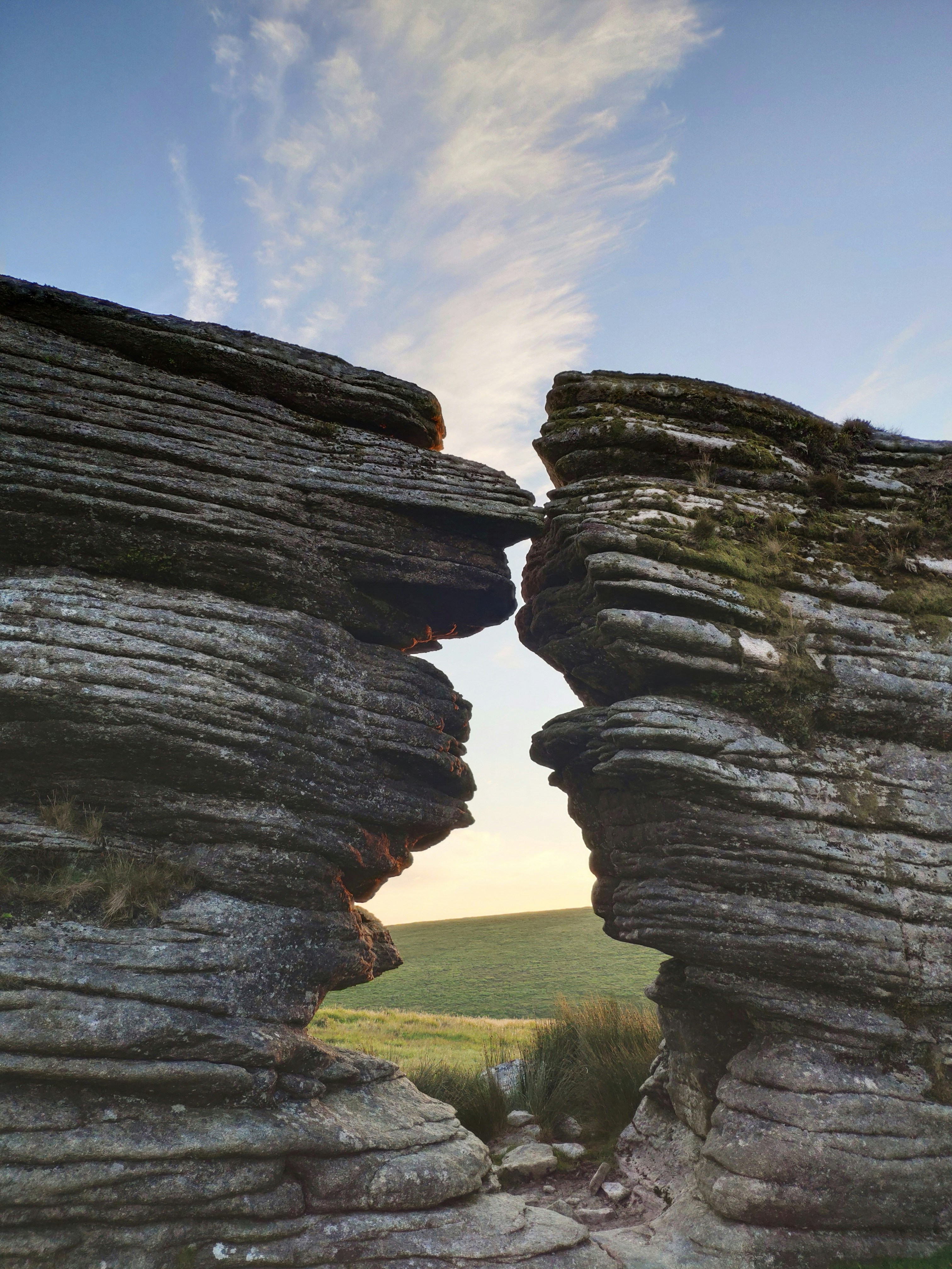 Two towering rock formations create a natural archway, framing the serene landscape beyond. The sky displays soft clouds as the sun sets.