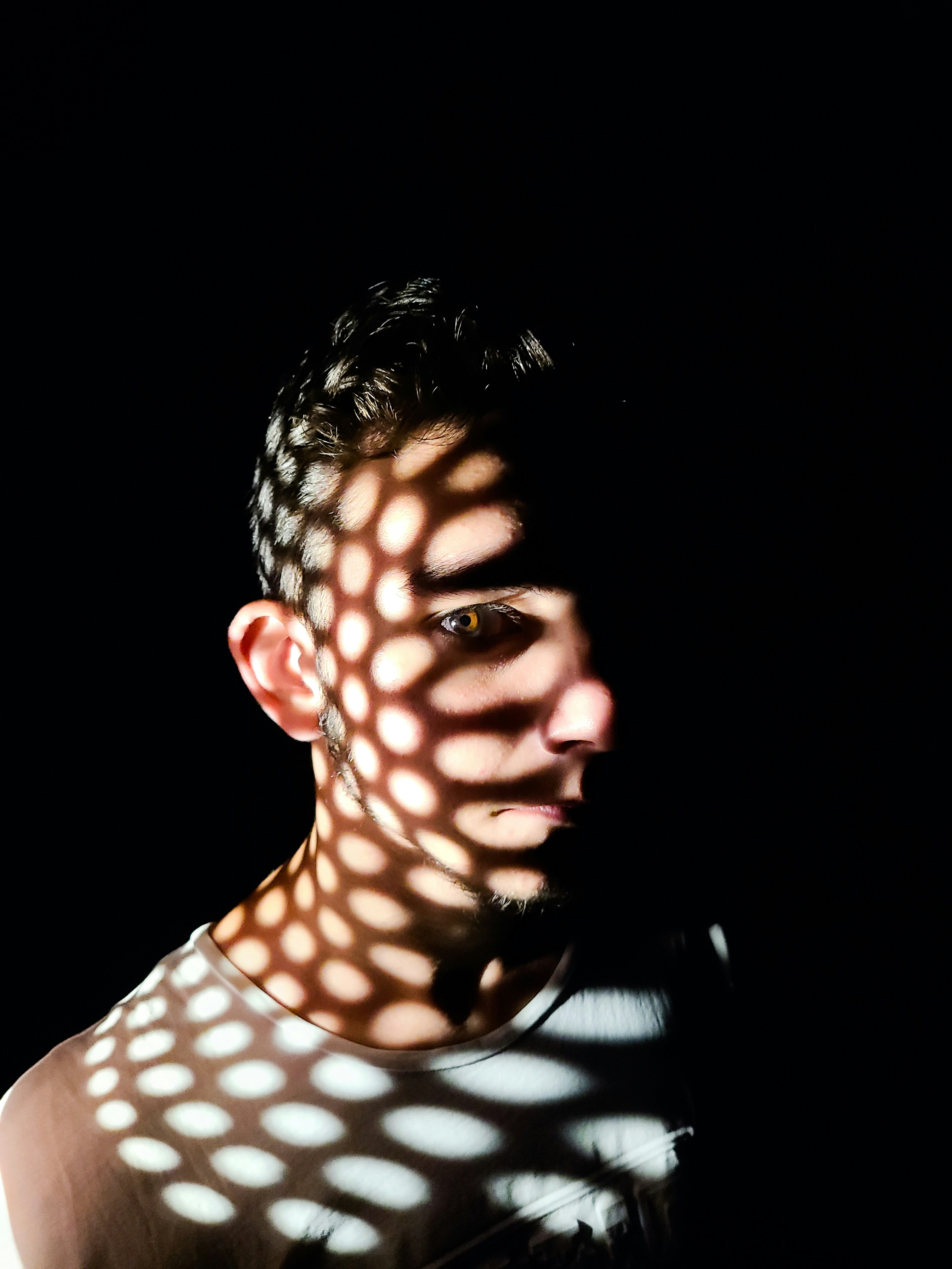 A portrait featuring a young man partially illuminated by intricate patterns of light and shadow. The interplay creates a dramatic and artistic effect.