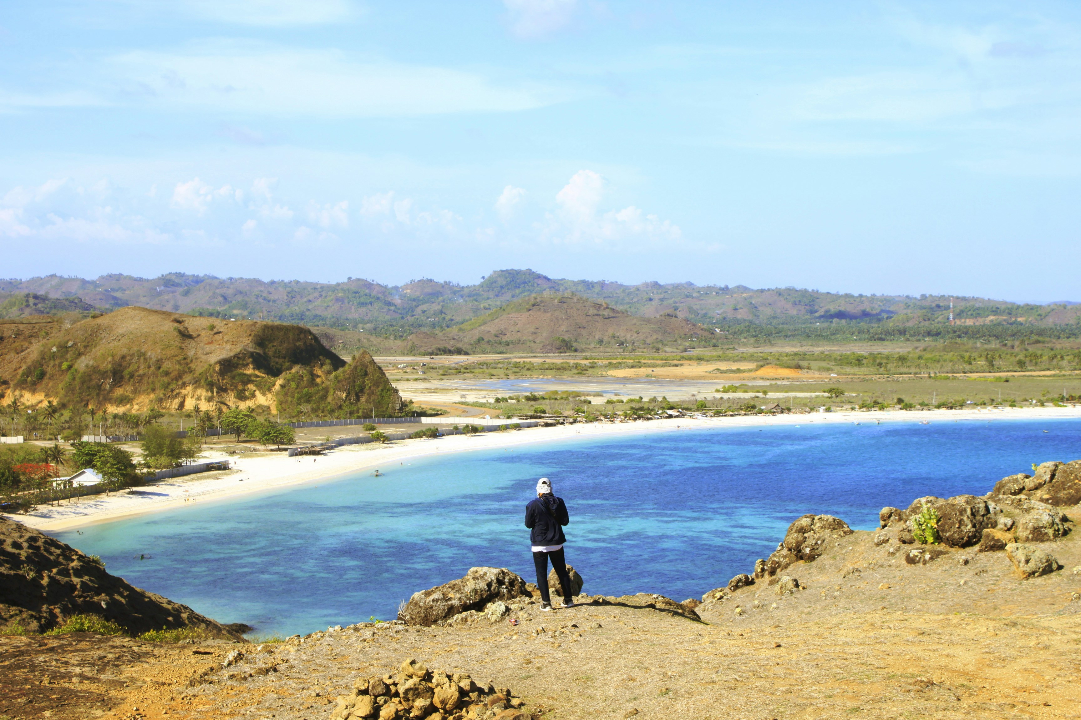 Person in a black jacket stands on a rocky cliff overlooking a vibrant blue ocean and distant hills in Indonesia.
