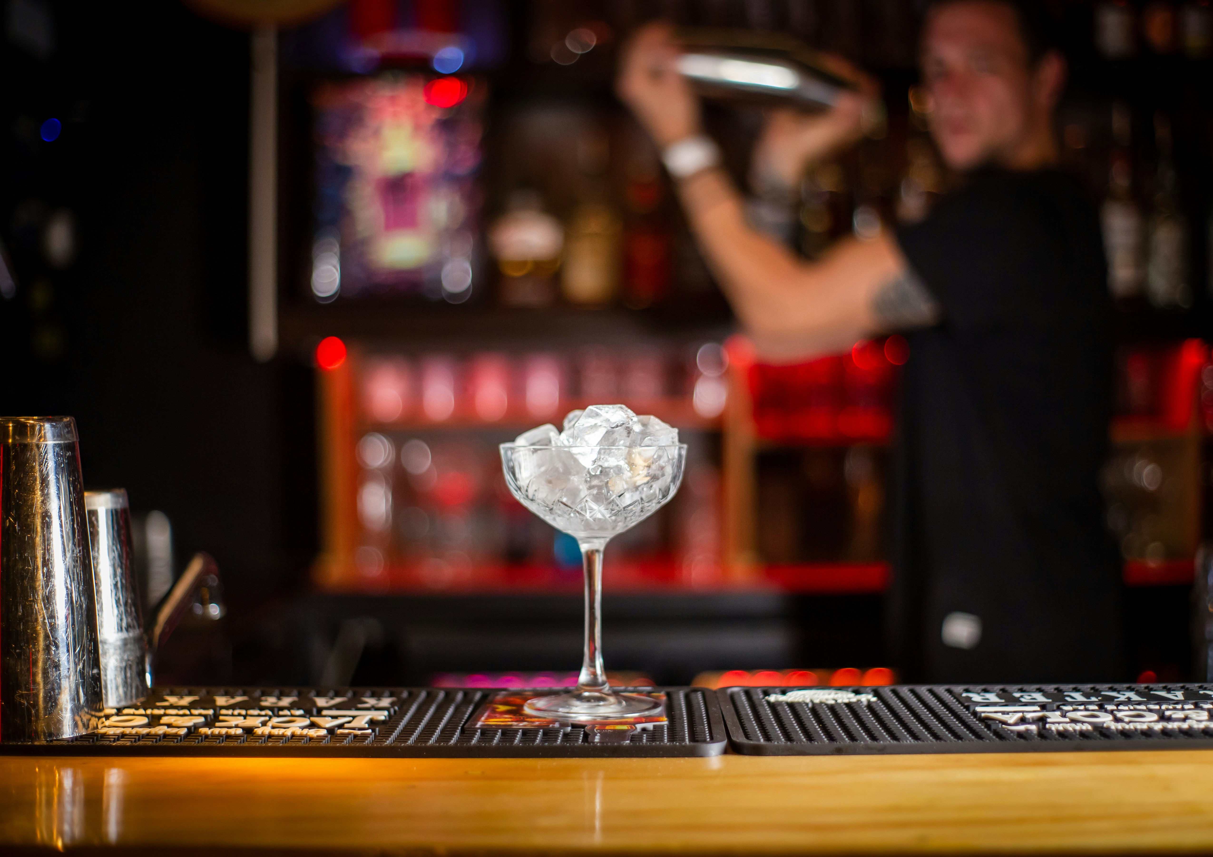 Cocktail glass filled with ice on a bar counter, with a bartender shaking a mixer in the background.