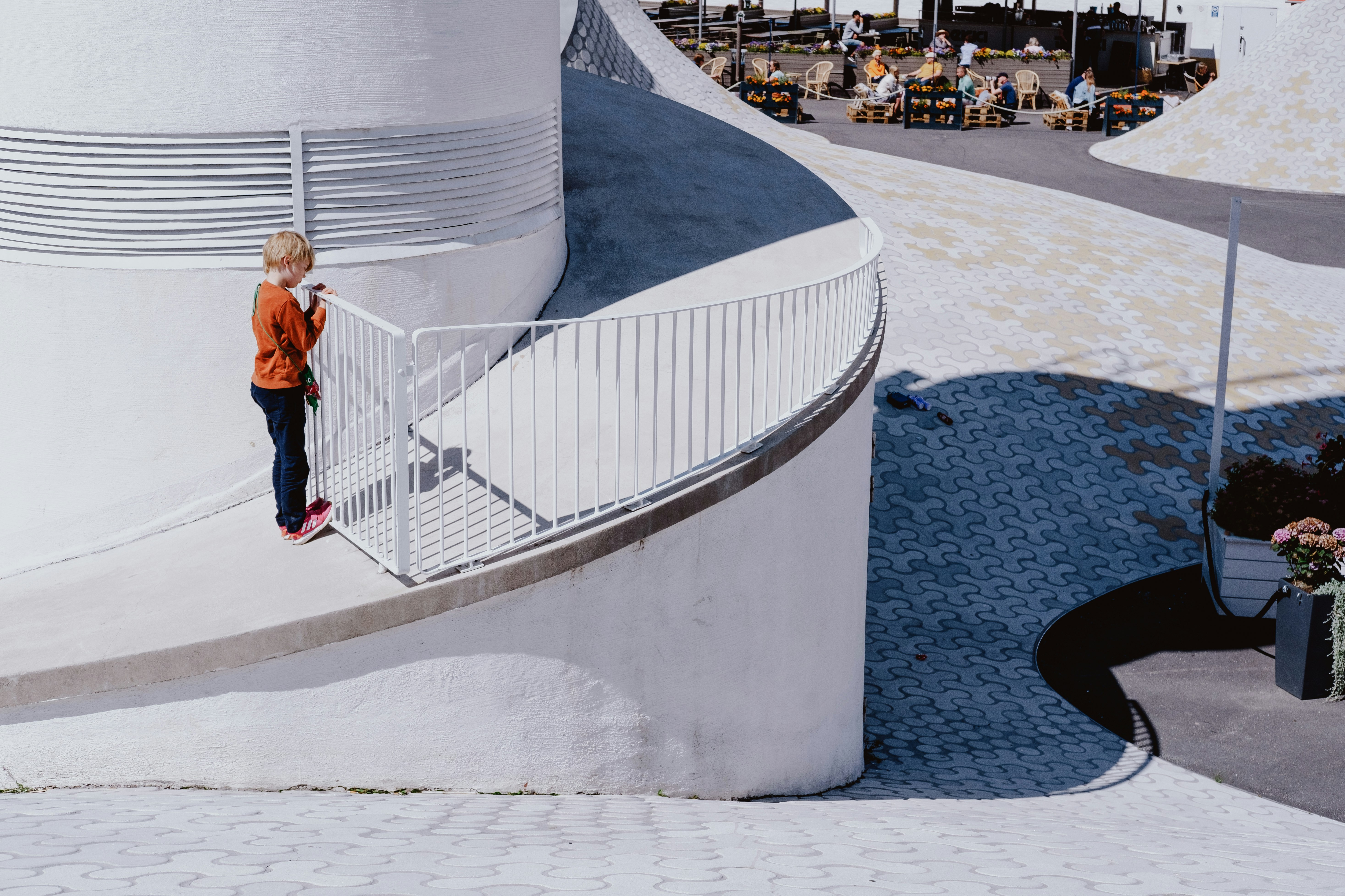 Child in an orange sweater stands on a curved ramp, observing the surroundings in a modern architectural space. The design features smooth lines and contrasting shadows.