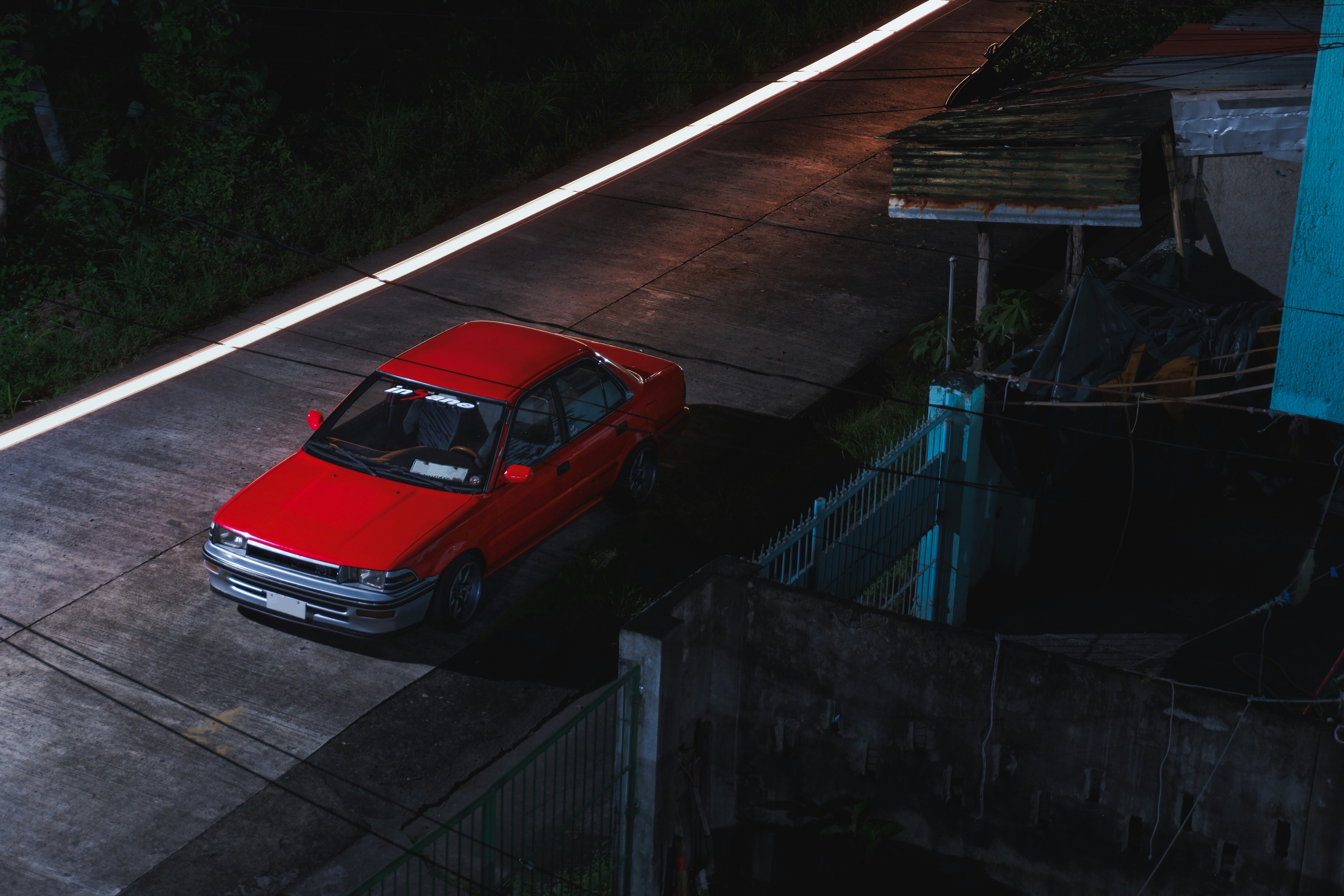 A vibrant red car parked beside a dimly lit road, with light trails illuminating the asphalt. The scene captures a blend of urban life and nighttime stillness.