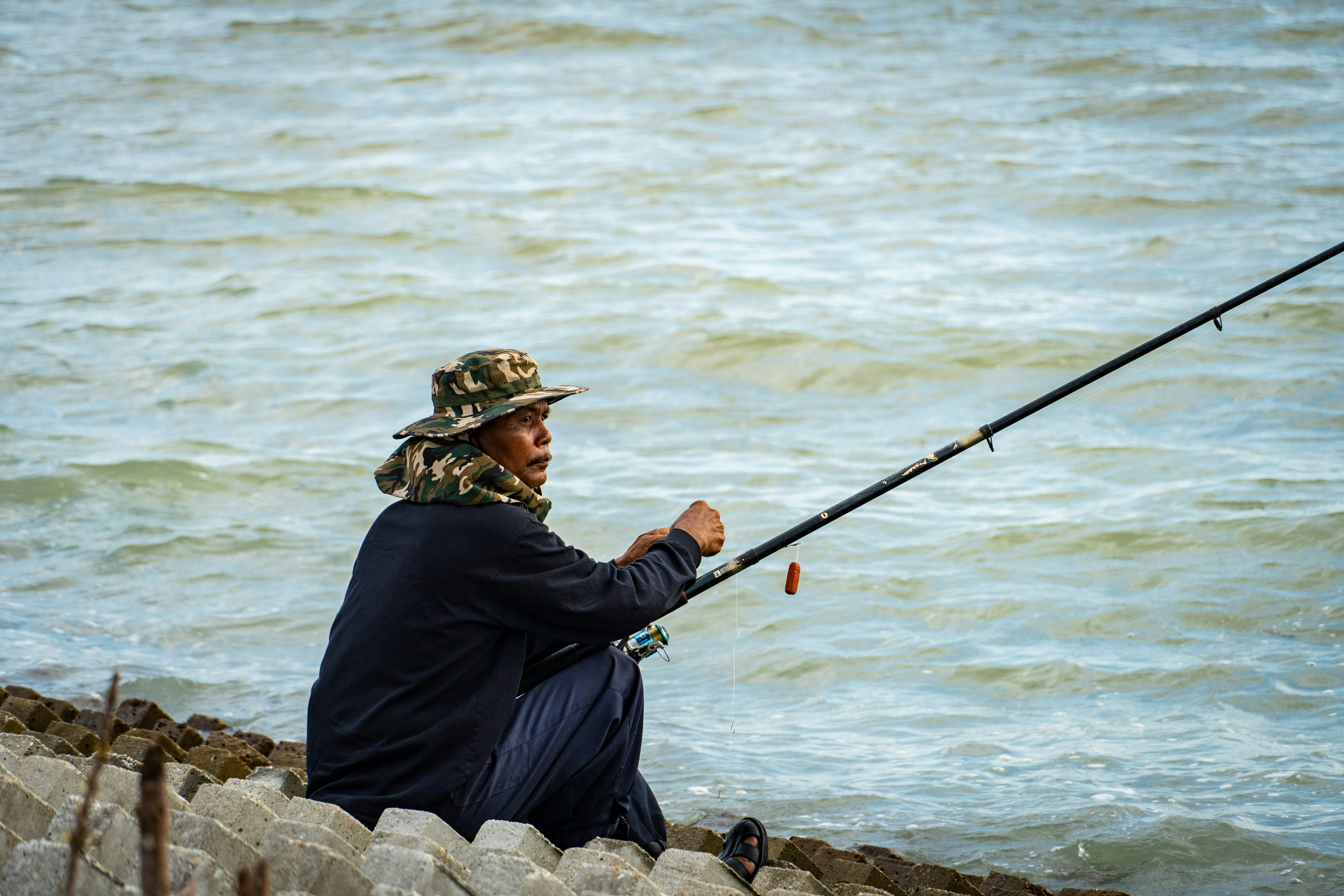 Fisherman seated on rocky shore, focused on his line against the backdrop of gentle waves. 