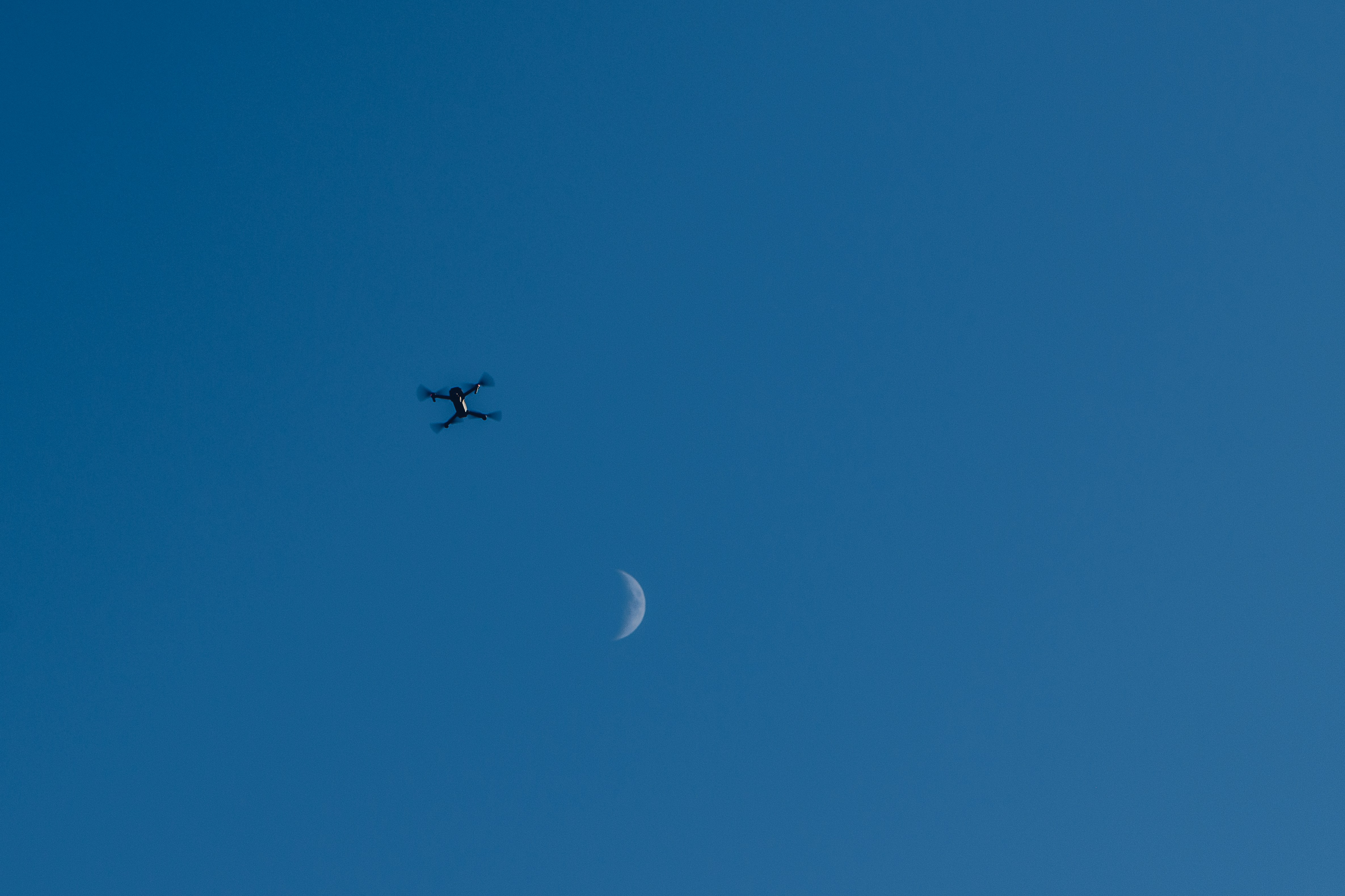 A drone gliding through a clear blue sky, juxtaposed against a crescent moon. The scene captures the intersection of technology and nature.