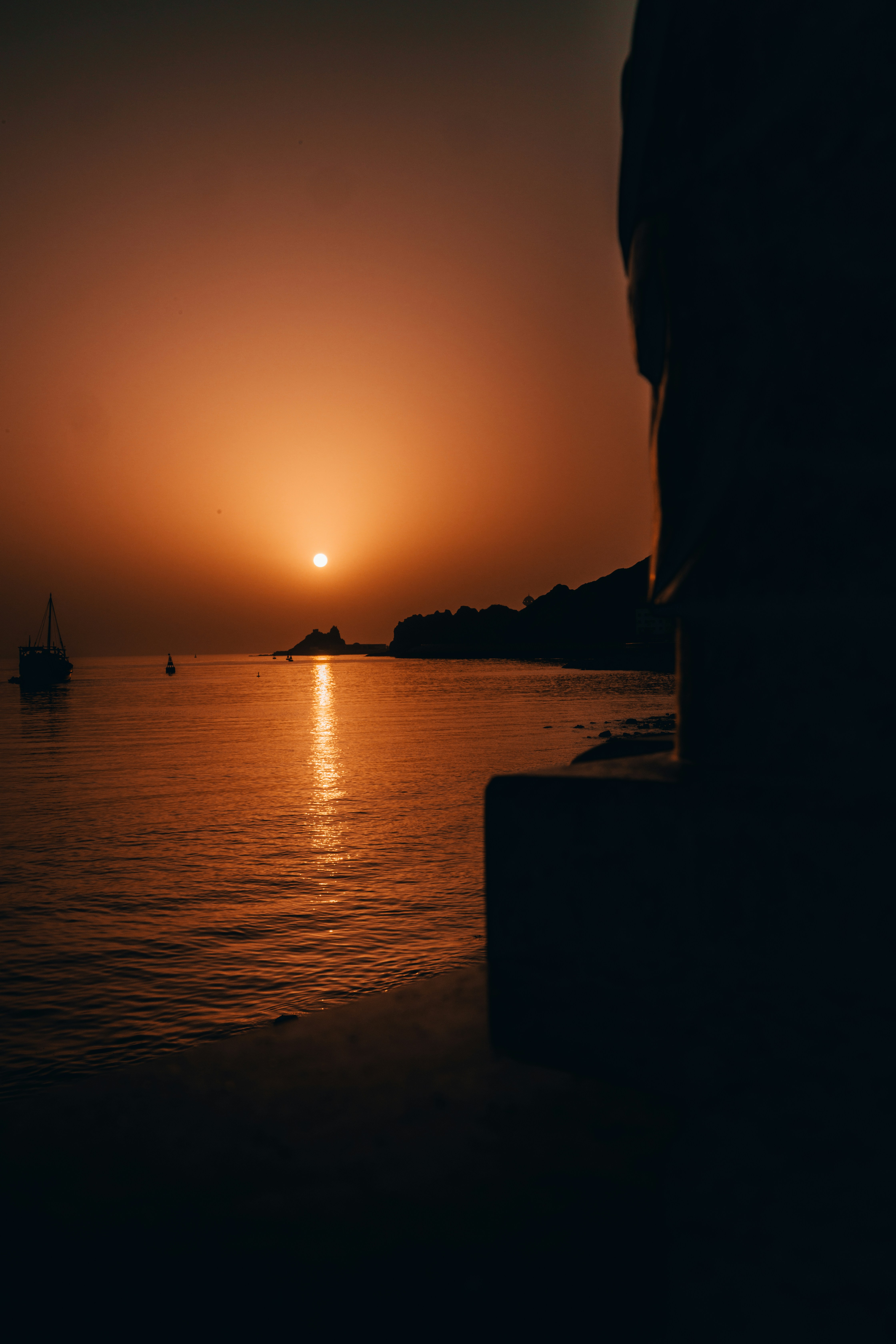 Silhouette of a statue beside a calm sea at sunset, with a glowing sun reflecting on the water's surface.