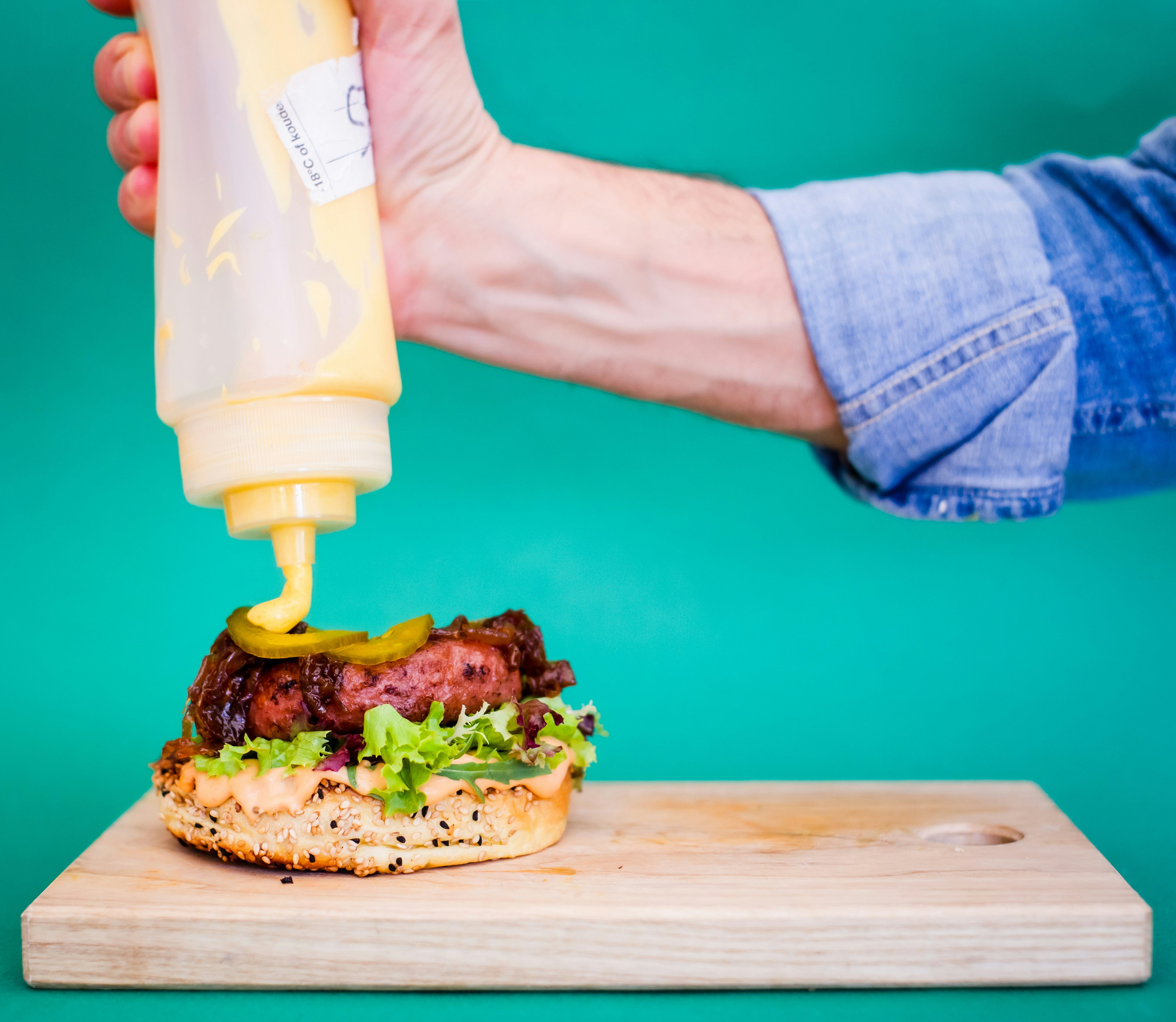 Hand squeezing mustard onto a gourmet burger with lettuce and beef patty on wooden board against teal background.