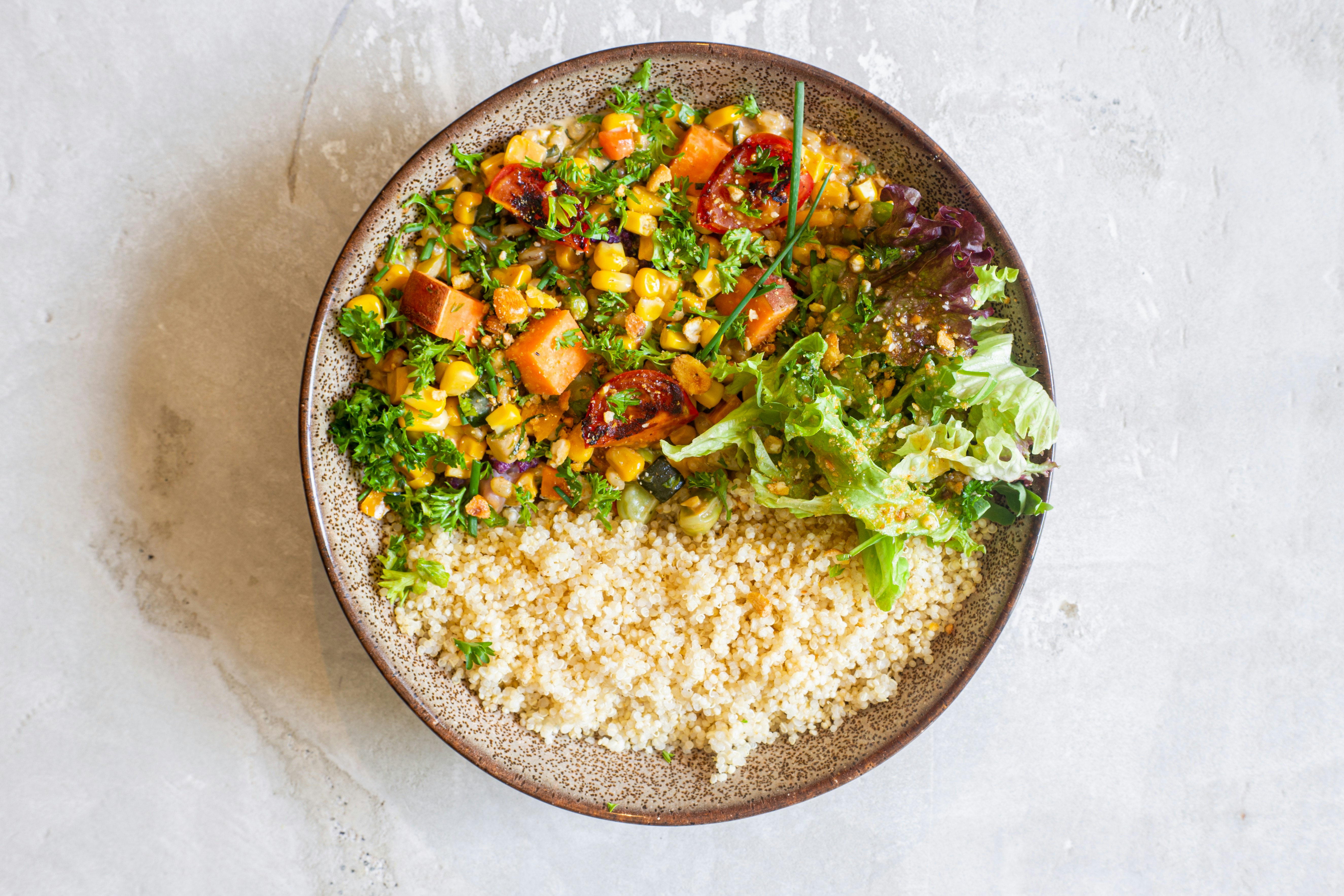 Colorful bowl of quinoa, roasted vegetables, corn, and fresh greens on a light background.