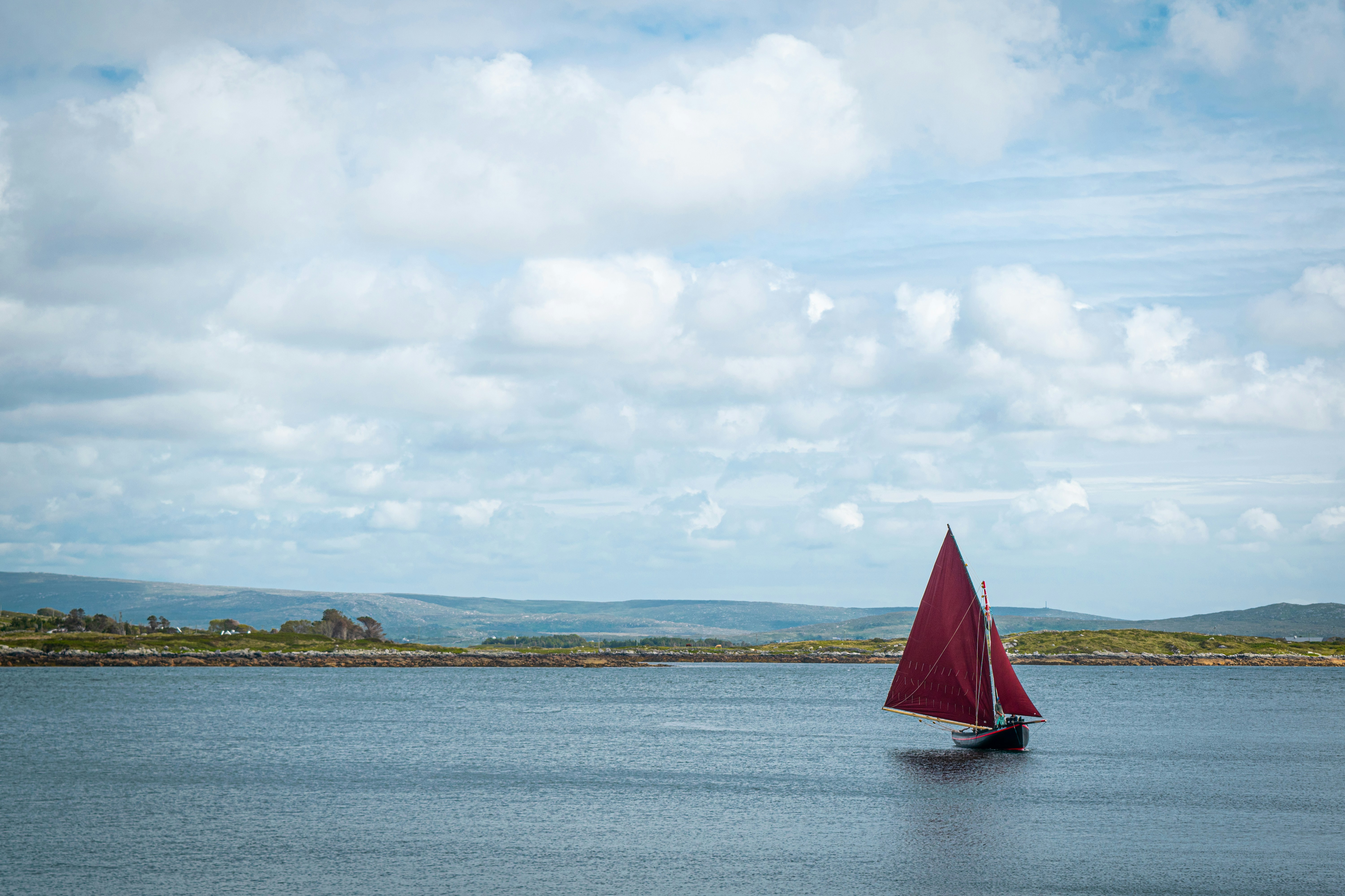 A sailboat with a vibrant red sail glides across a calm, expansive lake under a sky filled with soft clouds.
