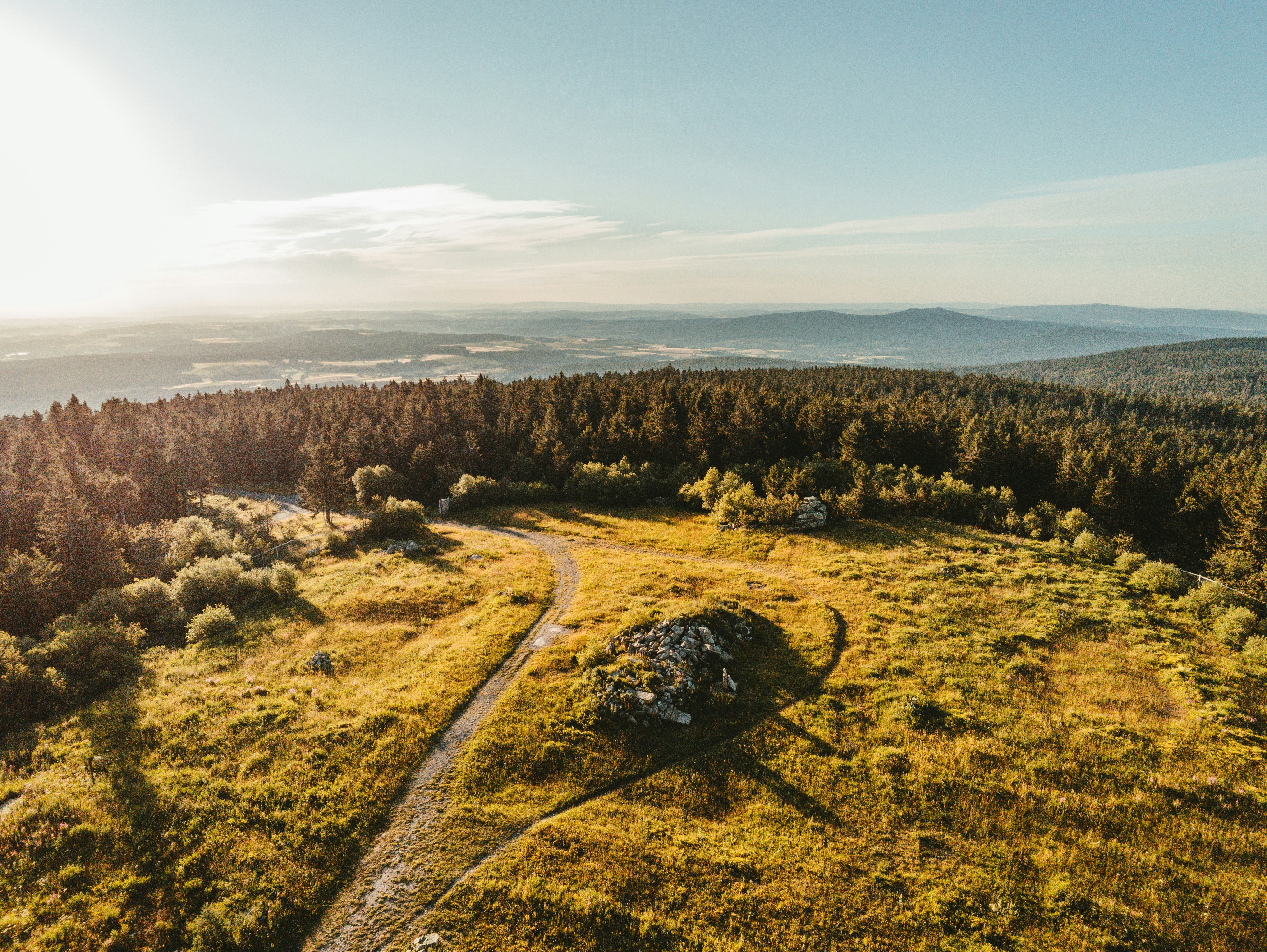 A peaceful meadow with a winding path and a large rock, surrounded by lush trees and rolling hills under a clear sky. The warm light of sunset casts gentle shadows across the landscape.