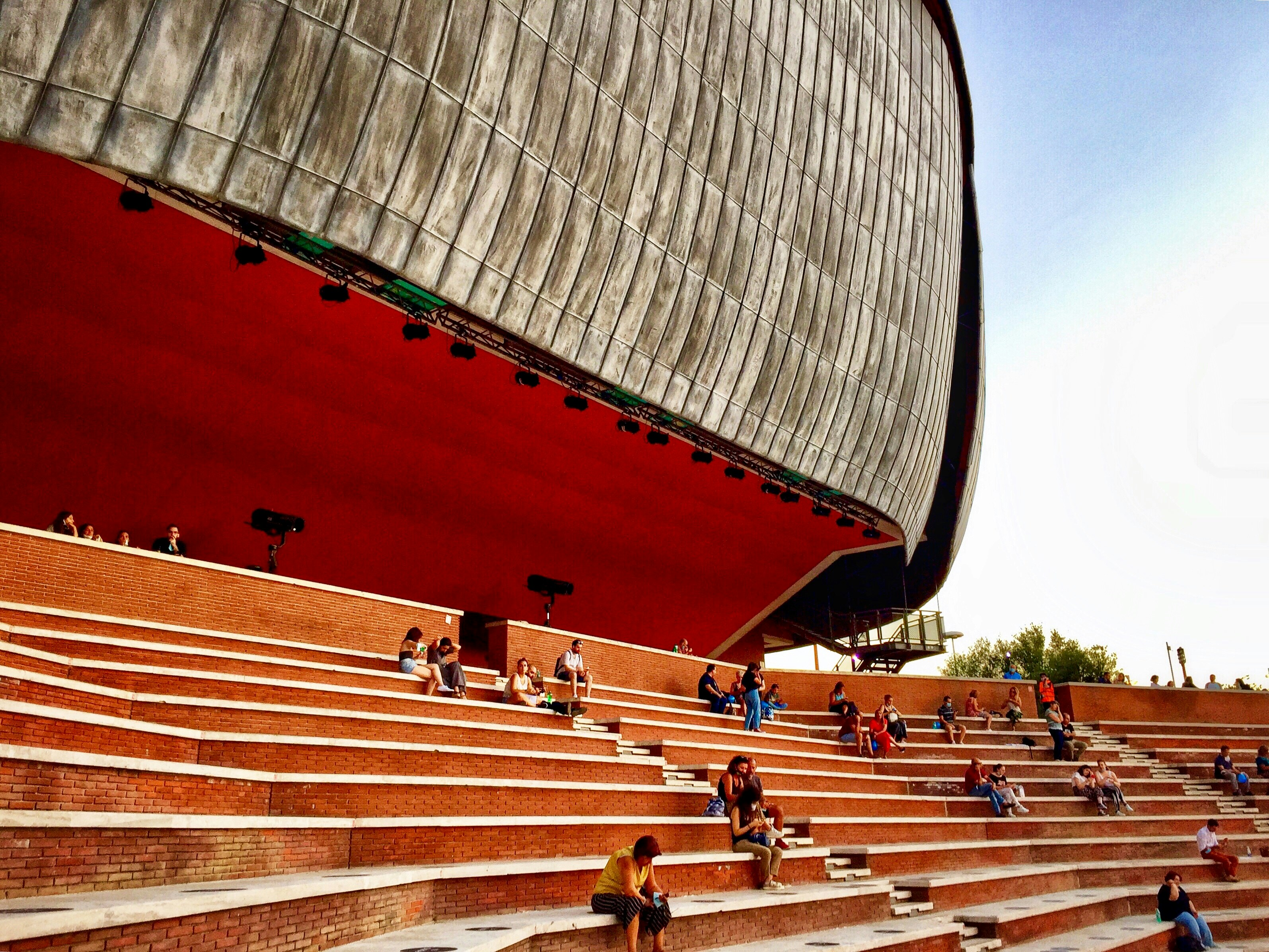 Modern amphitheater with a sweeping curved roof and tiered seating, filled with people enjoying the space.