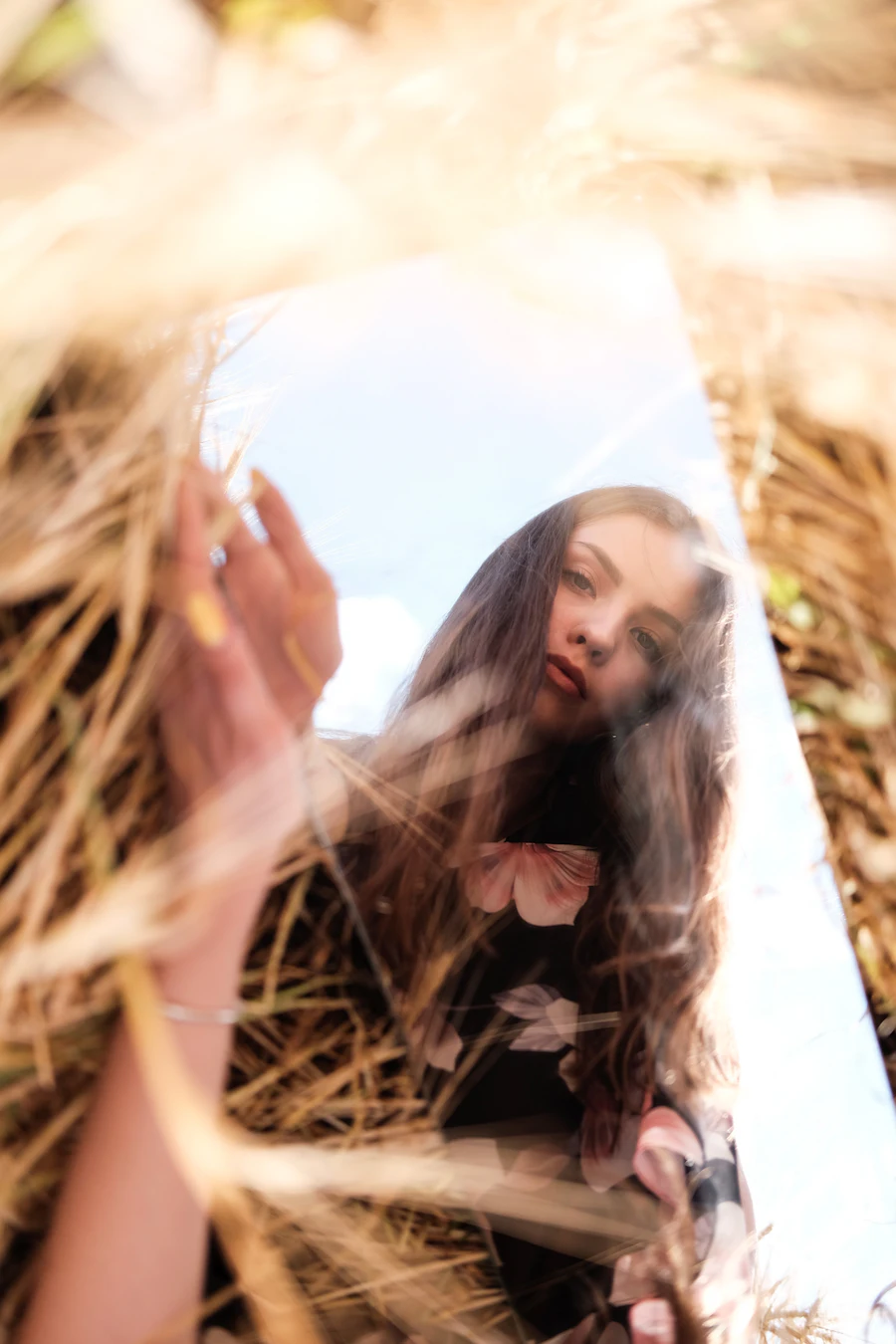 Portrait photographed through a mirror nestled in golden straw, with the subject’s face reflected against a pale blue sky.