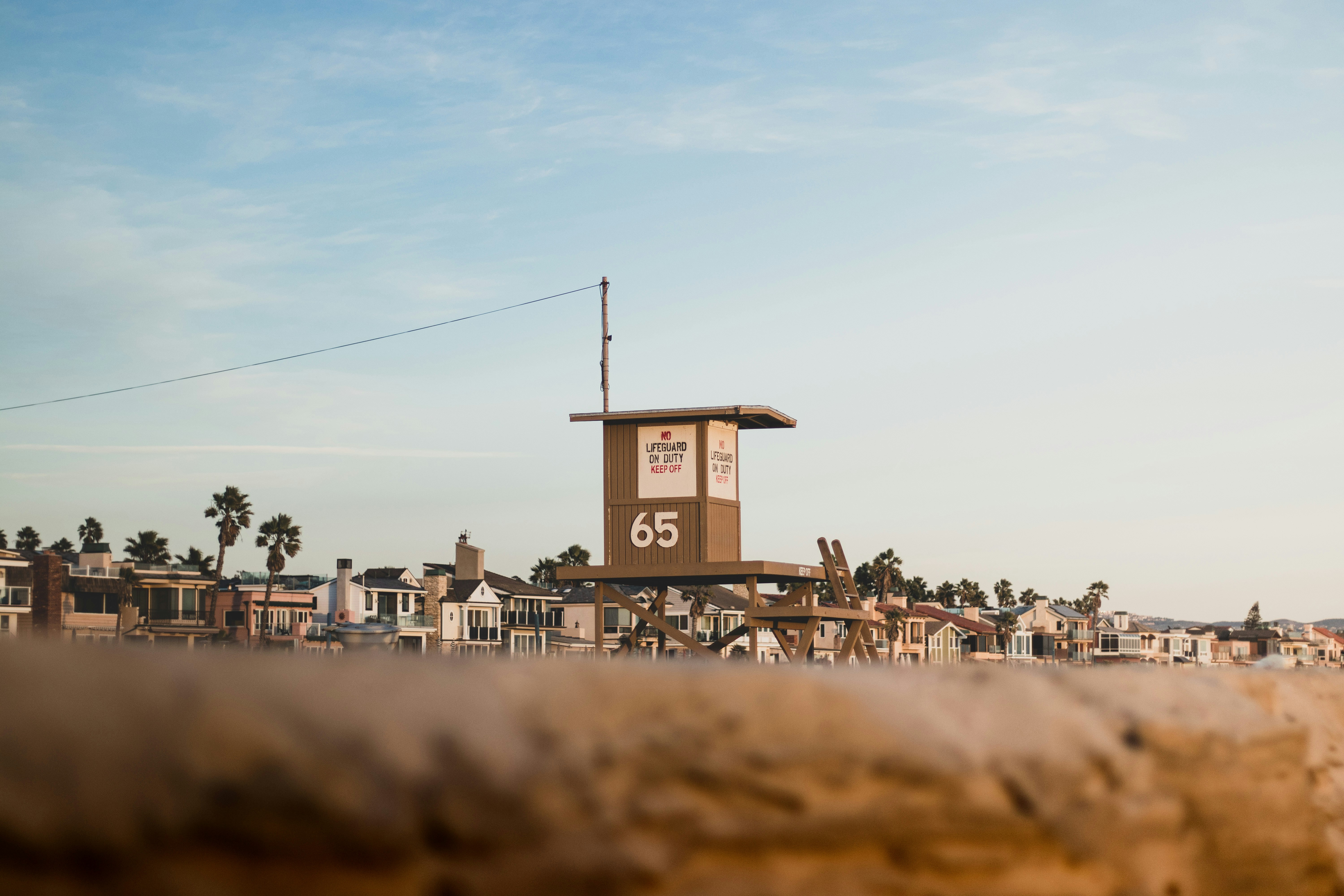 Lifeguard tower 65 overlooks a sandy beach as palm trees and houses line the background under a clear sky.