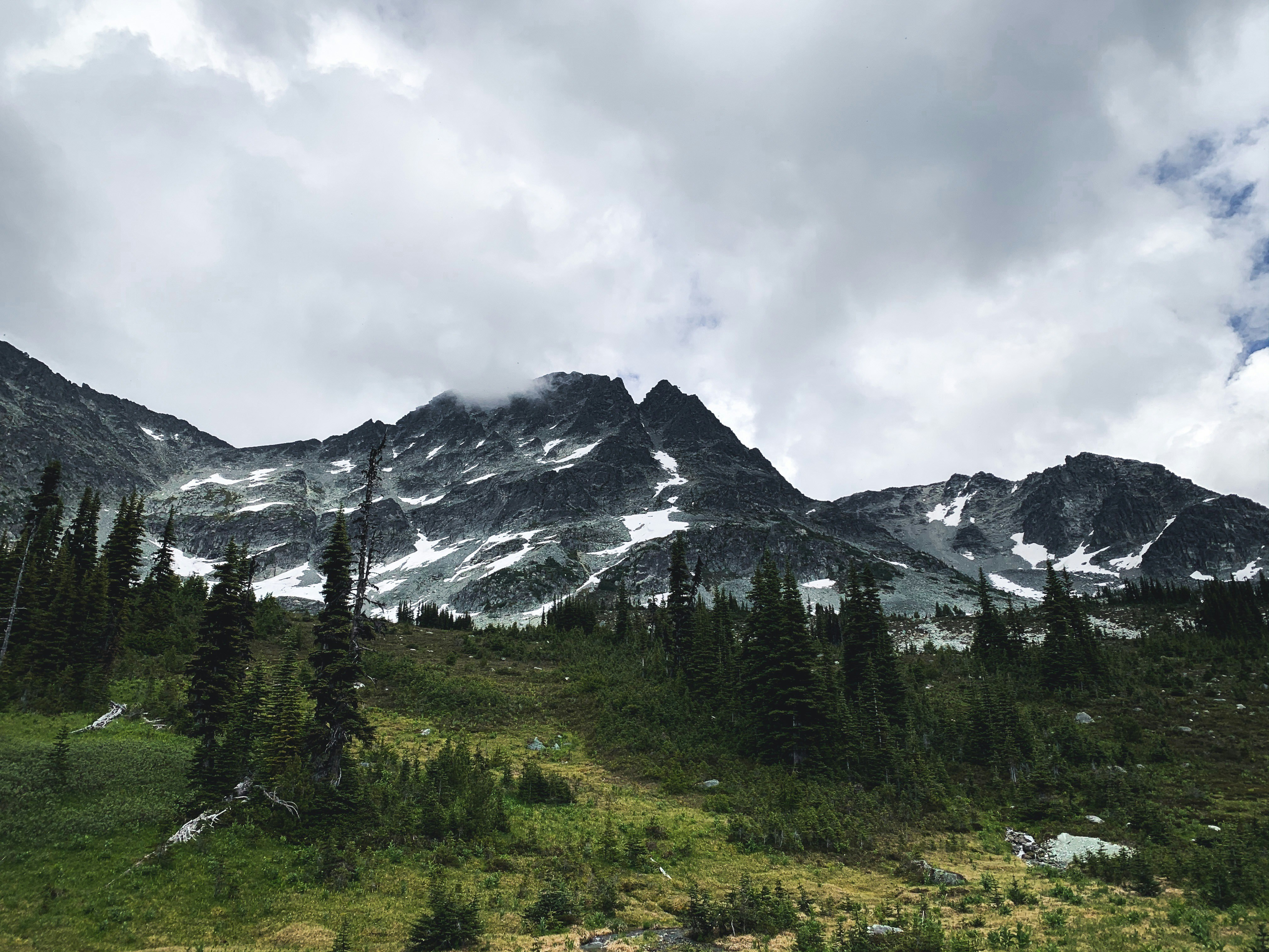 Dramatic mountain landscape with rugged peaks and patches of snow, framed by lush greenery and scattered trees. Clouds hover above, adding to the atmospheric depth.