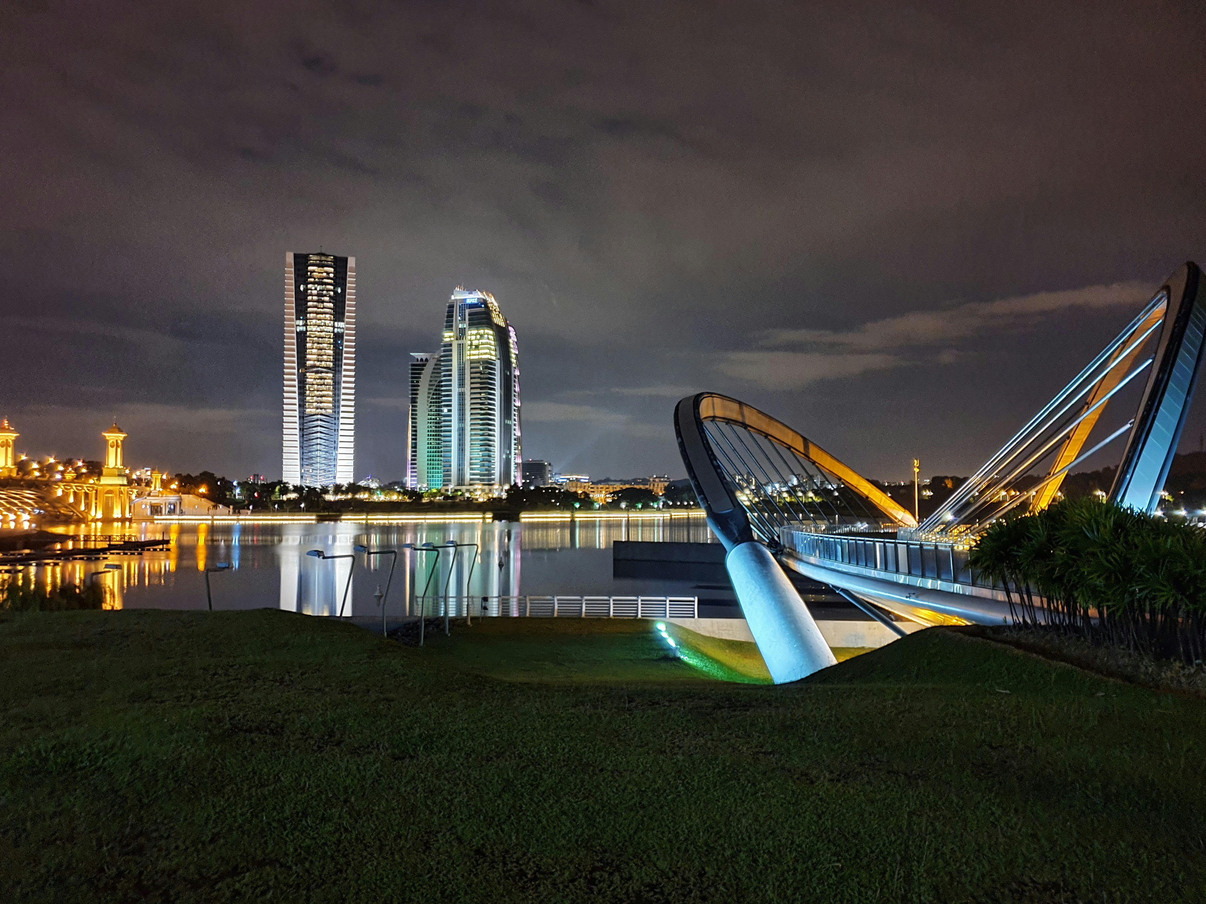 Illuminated skyline with modern architecture and a unique bridge reflecting in calm waters under a night sky.