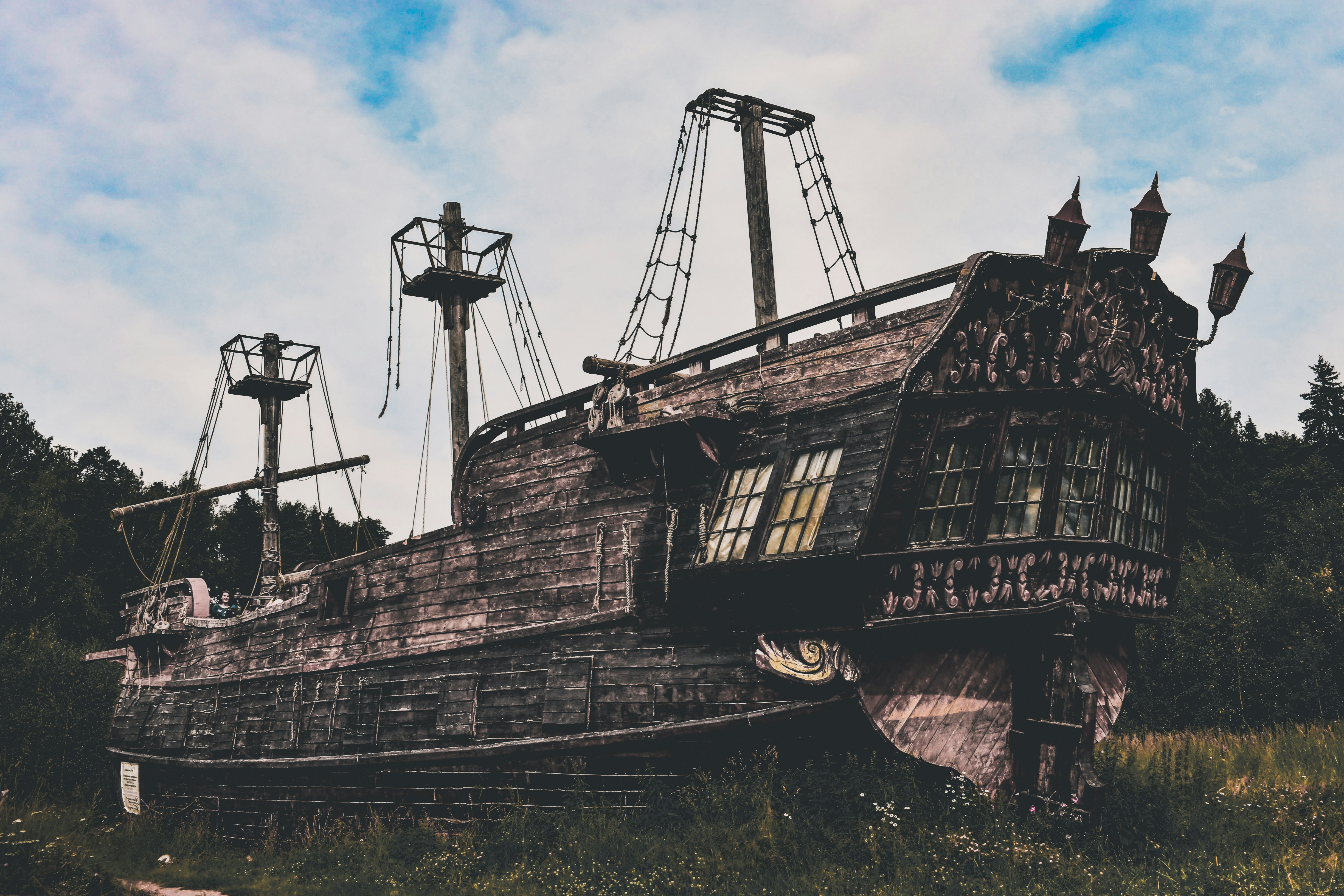 Abandoned wooden pirate ship resting on overgrown grass, showcasing intricate carvings and weathered sails against a cloudy sky.