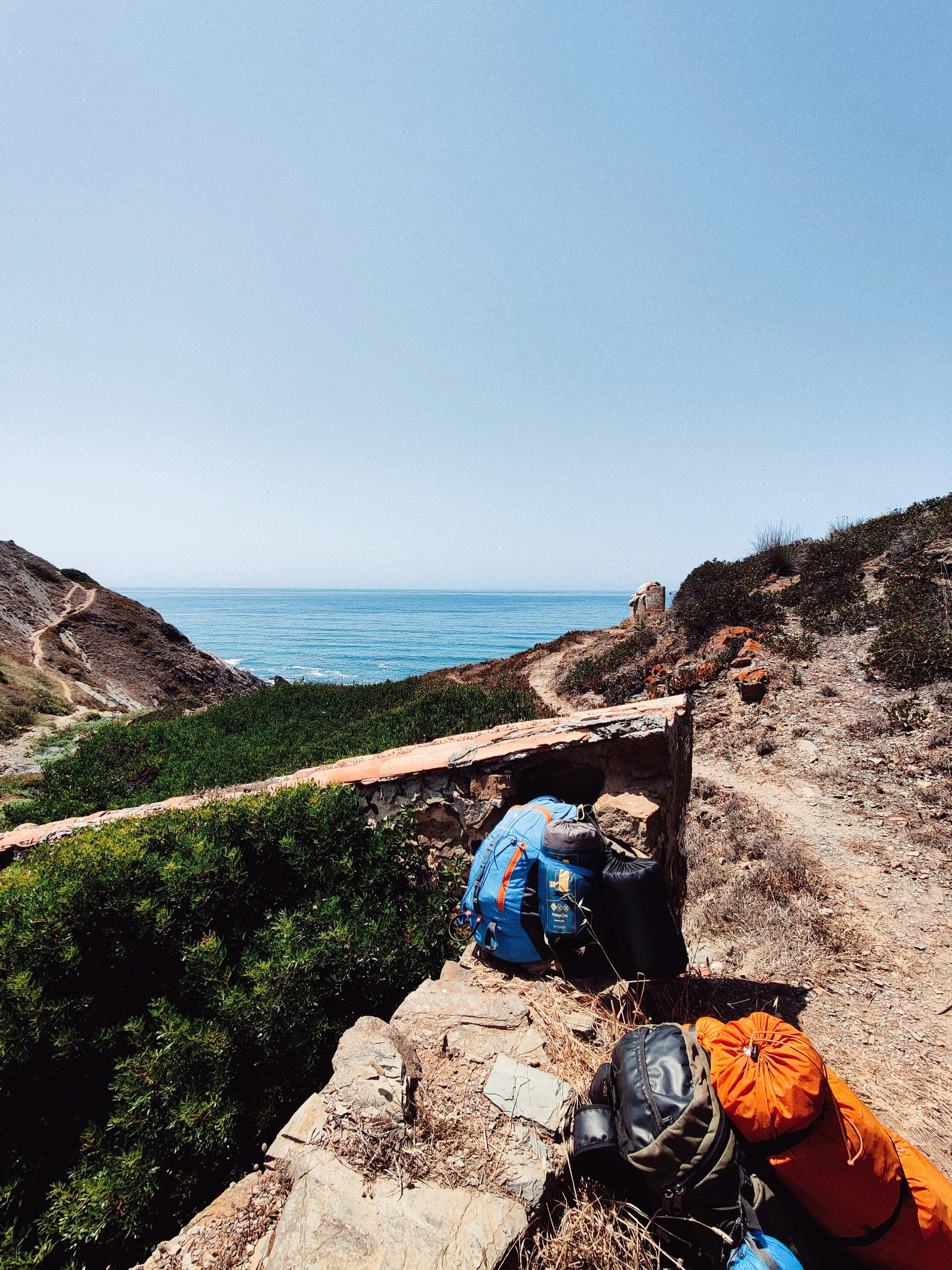 A rugged coastal trail scene with a blue backpack and other gear resting on sunlit rocks, overlooking the calm sea. The foreground gear anchors the composition against scrubby dunes and a clear sky.