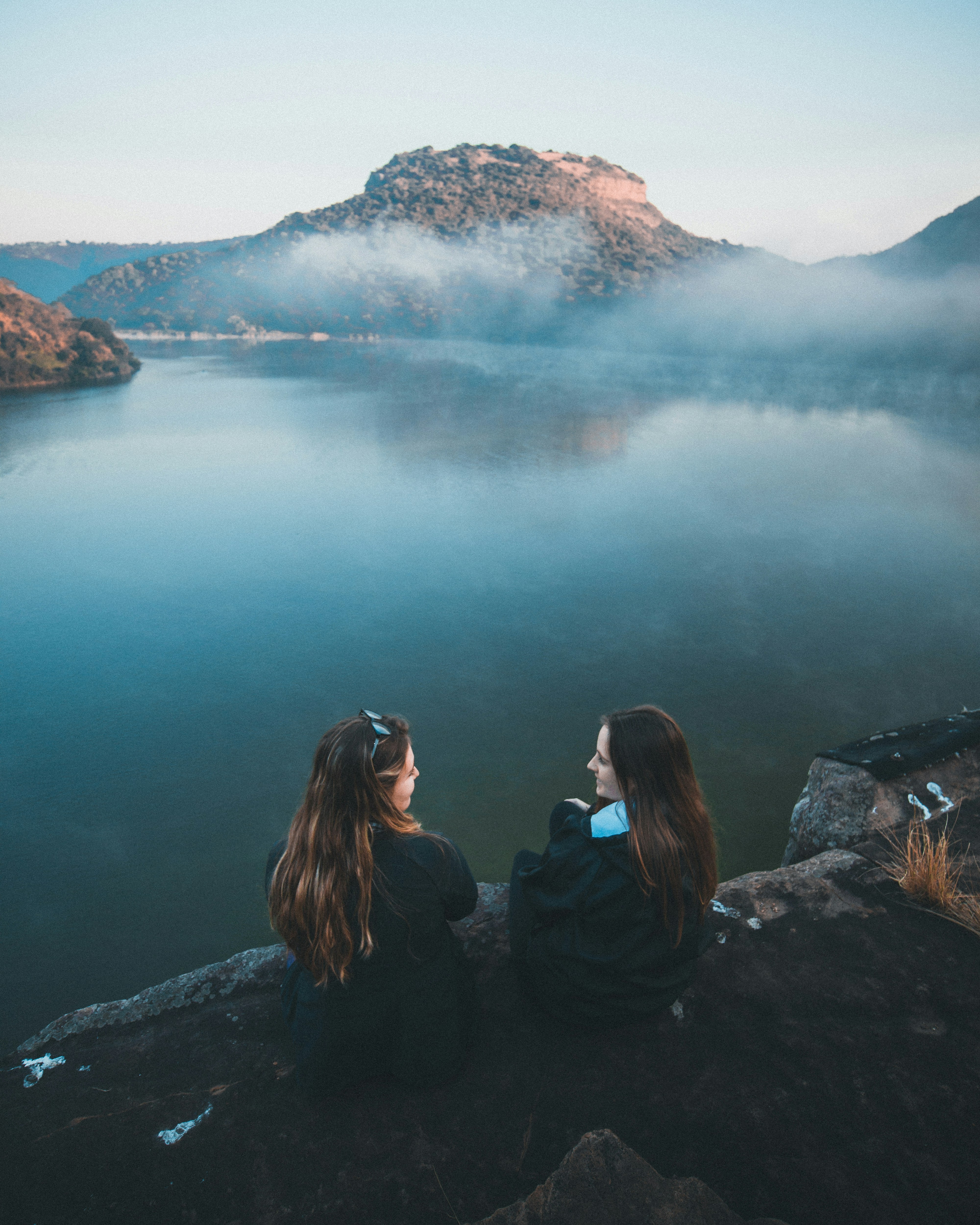 Two friends sitting on a rocky ledge overlooking a tranquil lake surrounded by misty mountains. The calm water reflects the serene landscape.