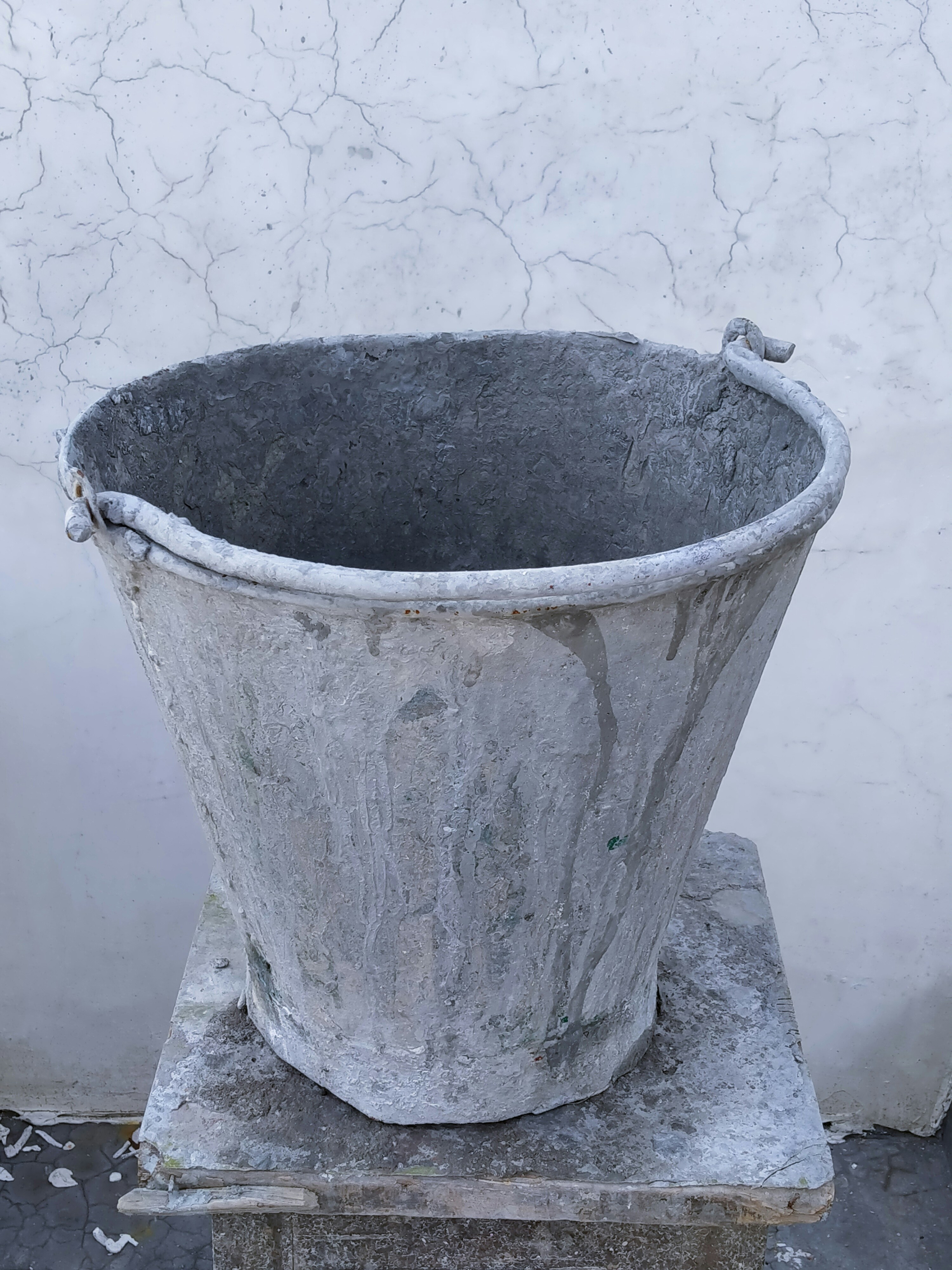 A weathered concrete bucket with visible wear and tear, resting on a stone pedestal against a cracked white wall.