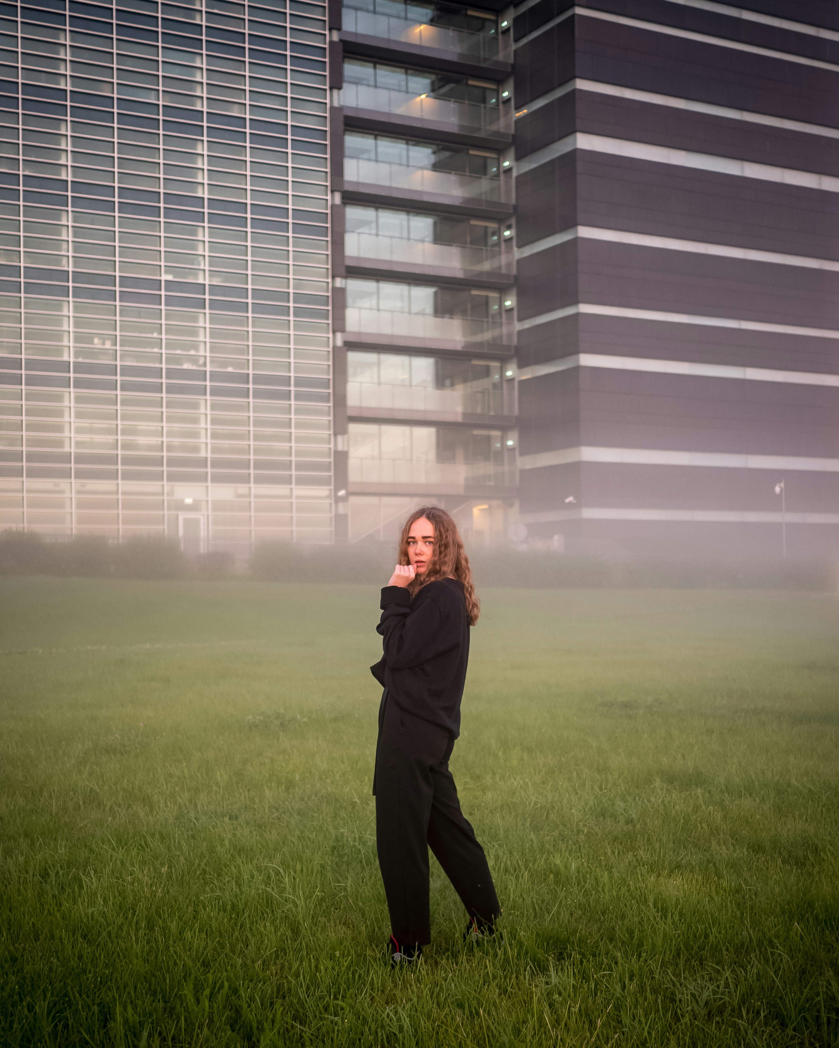 A young woman stands in a misty field, framed by a modern glass building, exuding a contemplative aura. The ethereal fog enhances the serene atmosphere.