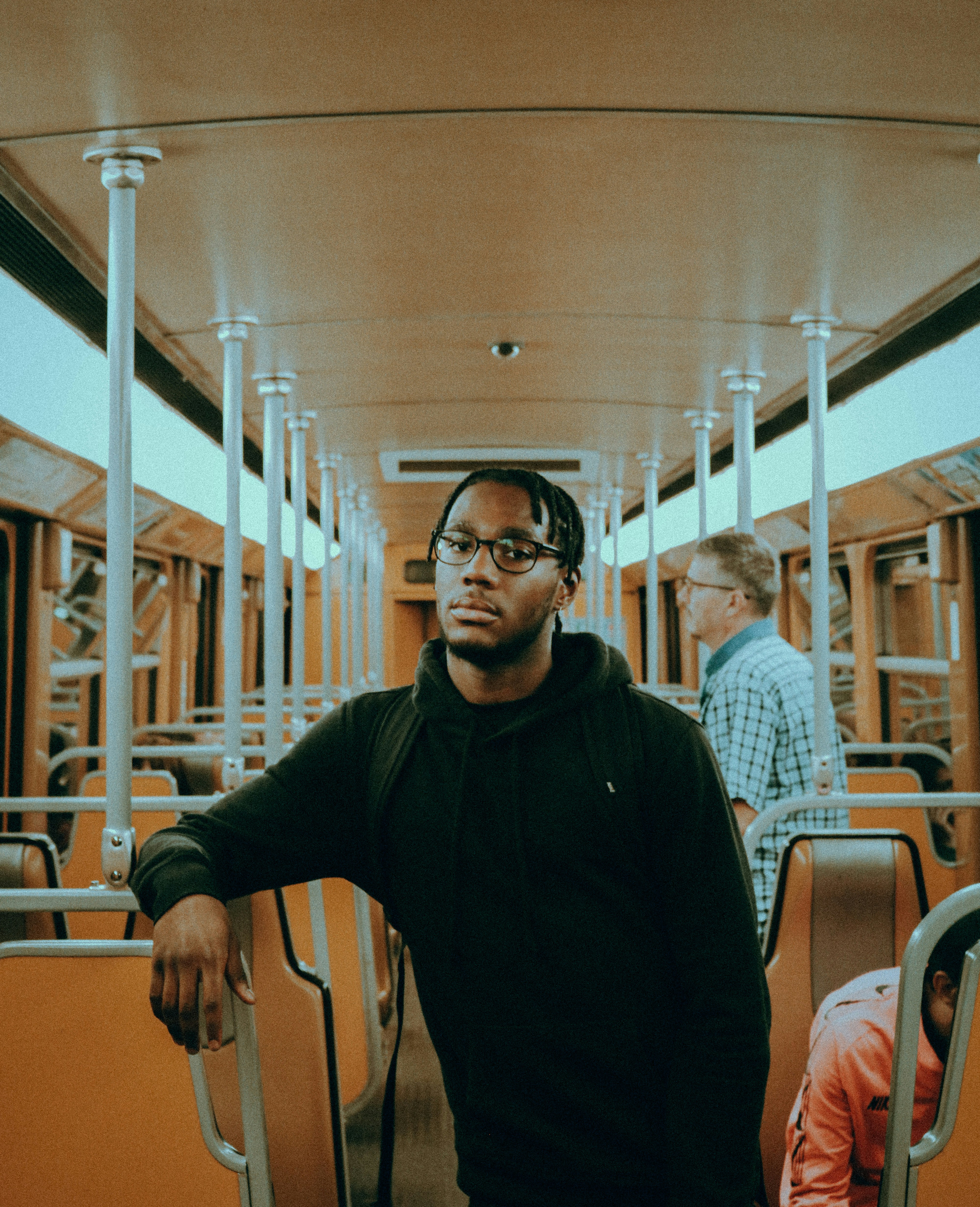 A young man in a black hoodie stands confidently in a nearly empty train, surrounded by warm orange tones and soft lighting. The atmosphere conveys a sense of introspection amidst urban travel.