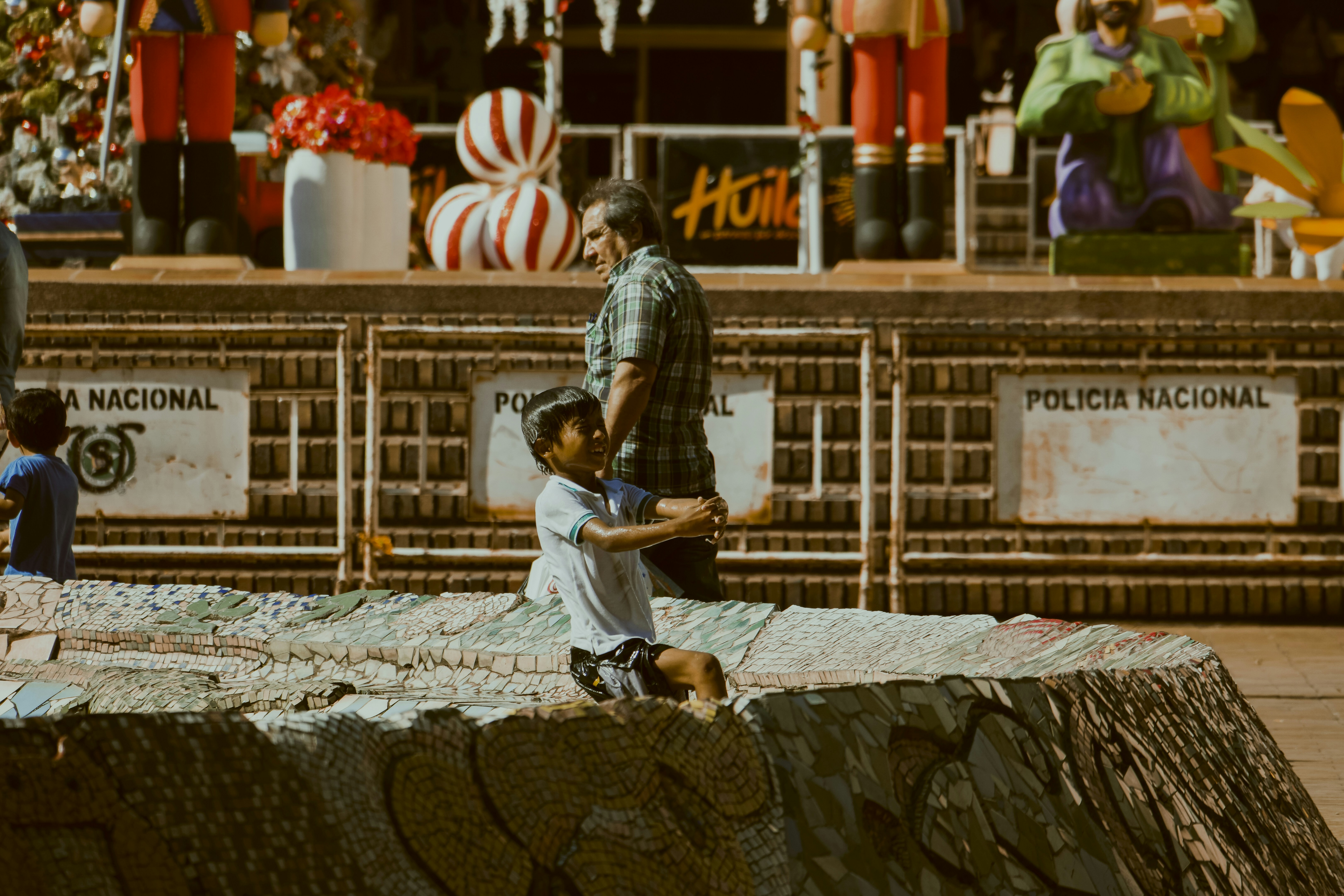 A young boy plays joyfully on a colorful mosaic while an adult observes in a lively public square adorned with festive decorations.